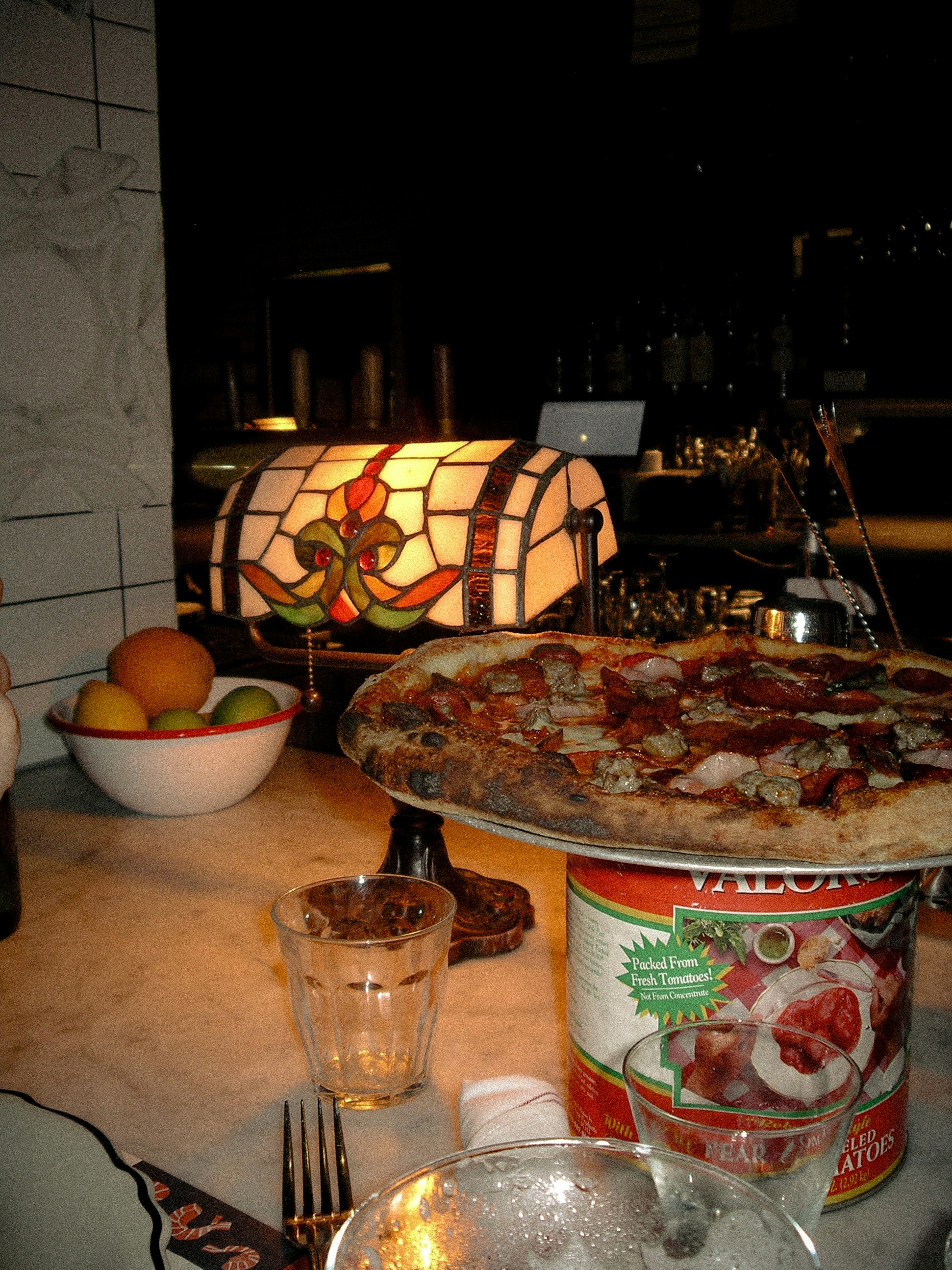 A pizza on a tomato can beside a stained glass lamp and fruit bowl.