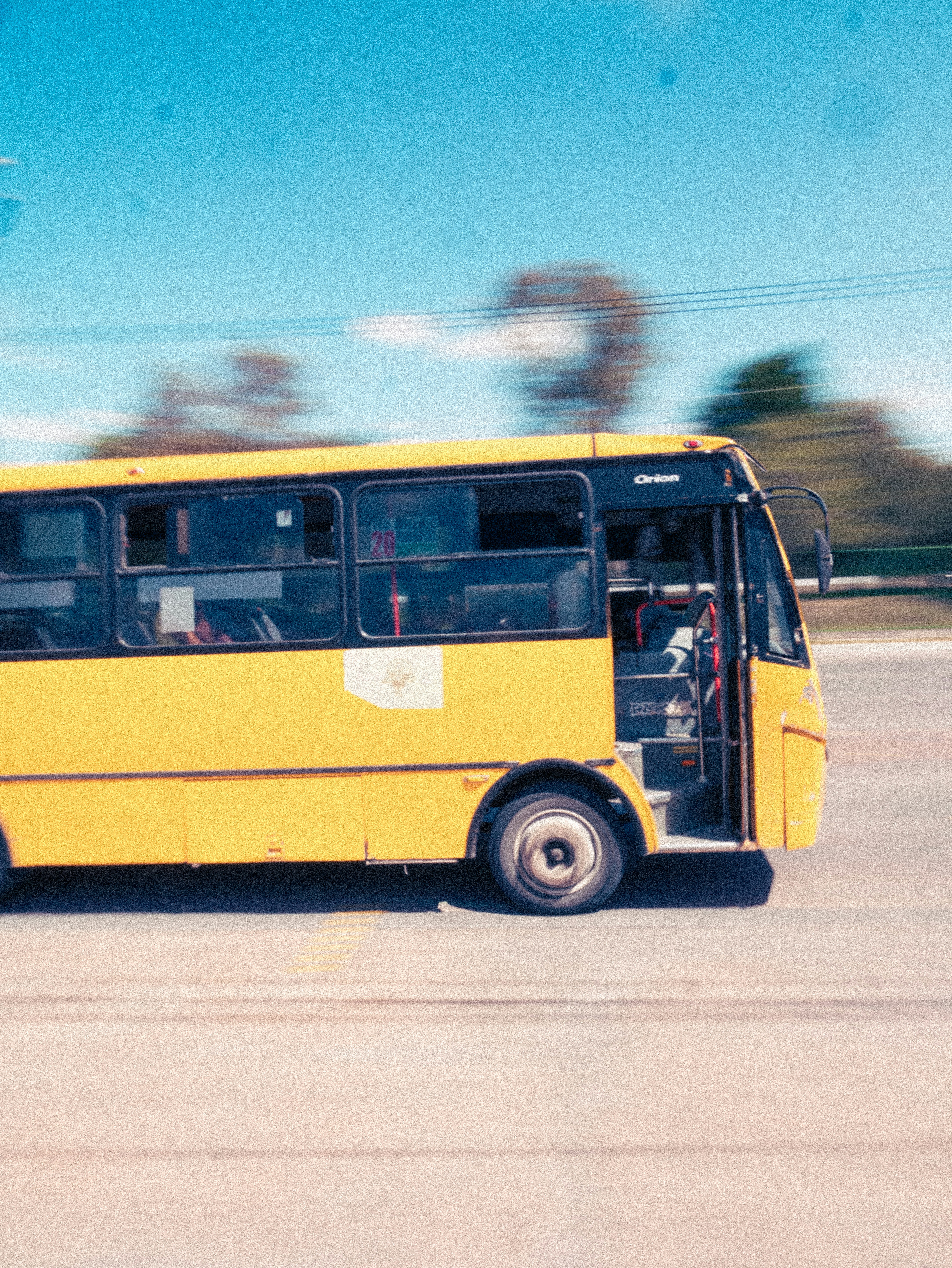 A yellow school bus rolls past in motion, with motion blur emphasizing speed in an open parking lot. The vehicle dominates the frame, showing an open door and windows, while a pale blue sky and distant trees provide a backdrop. Logo or branding appears on the bus side.