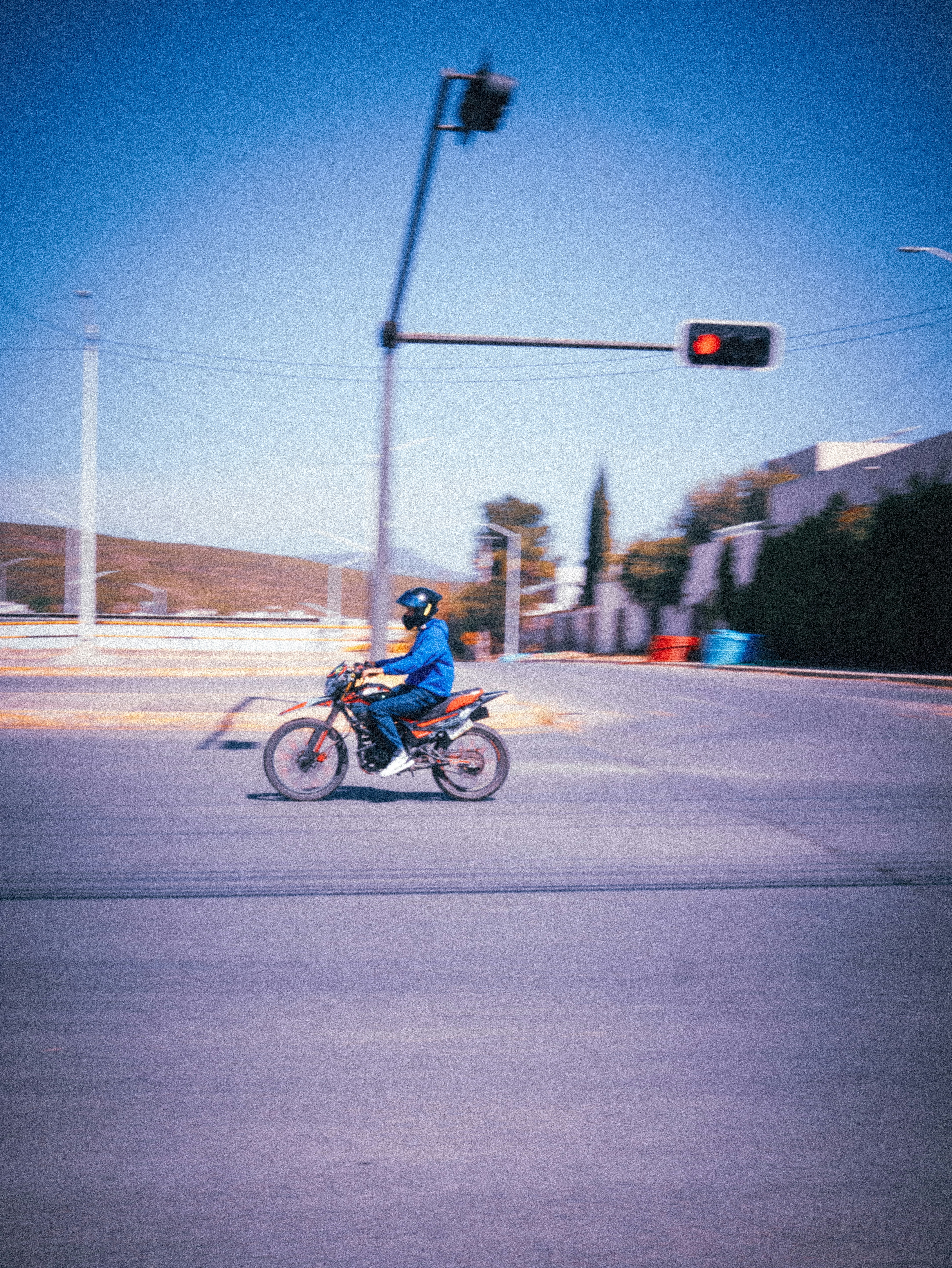 Blue-jacket motorcyclist rides through an intersection with a blurred, grainy look; street, traffic light, and distant buildings provide context. Note: image appears as a textured photograph.