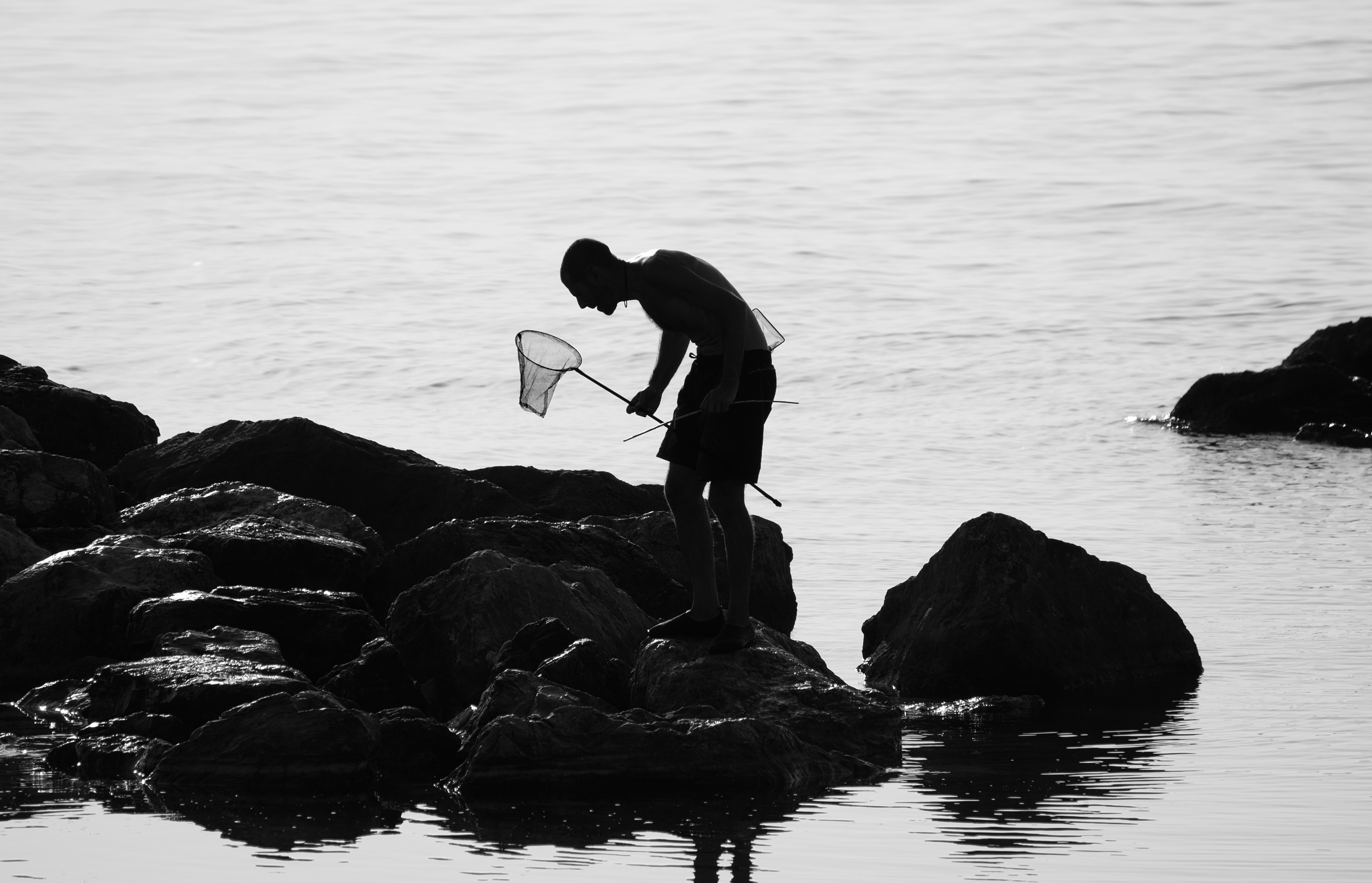 Monochrome scene of a silhouetted netslinger perched on rocks near calm water, catching light with the catching net as the tide recedes.