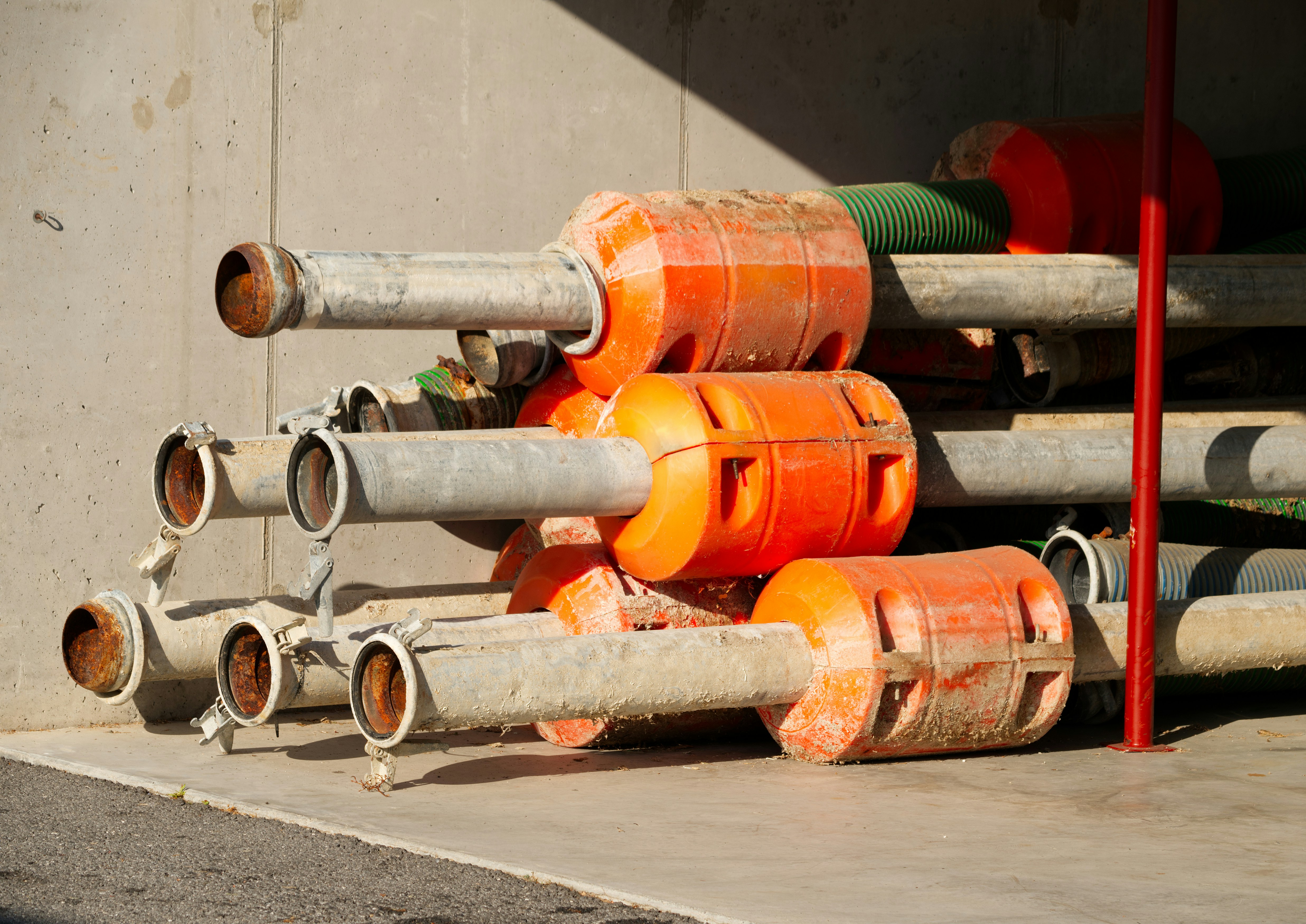 Stacked orange-capped pipes and metal tubes lie near a concrete wall, with a red vertical post in the scene, highlighting a still moment at a construction site.