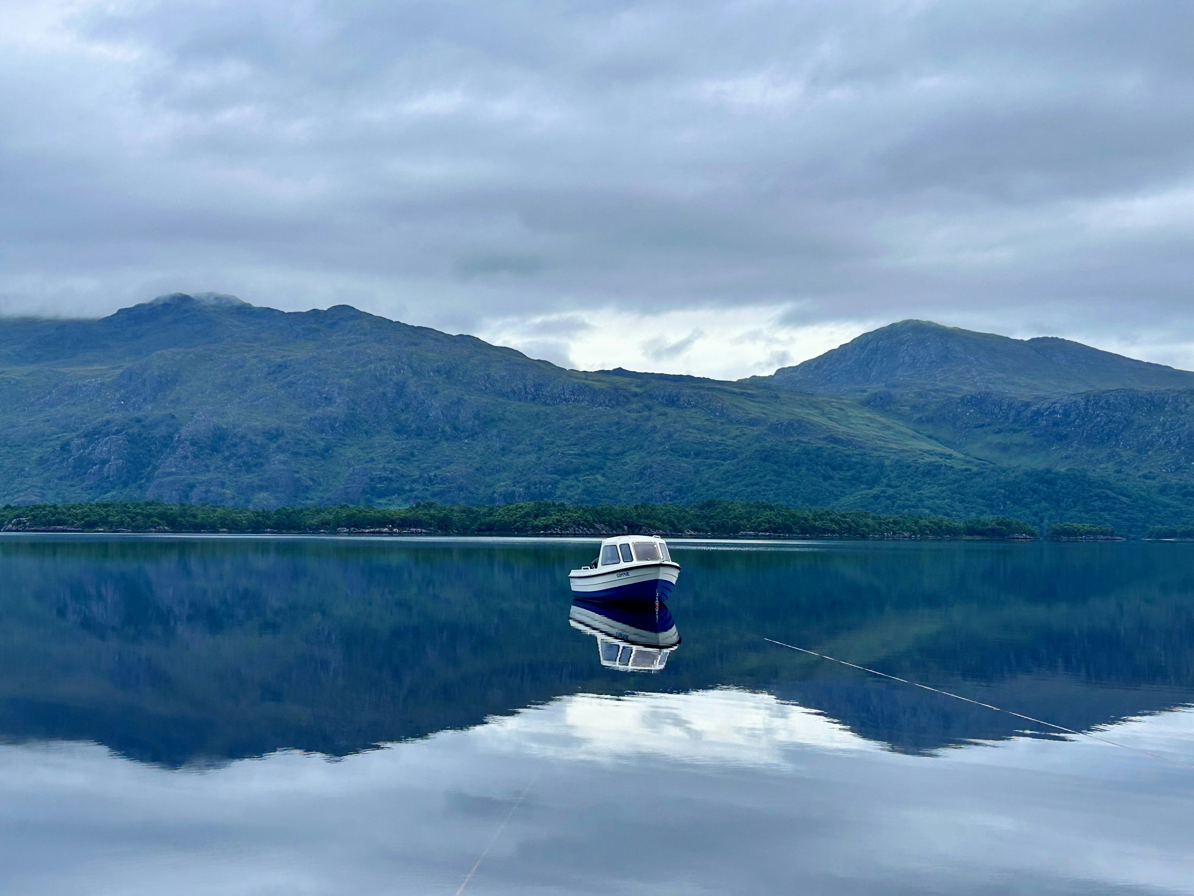 Small motorboat rests on glassy lake with distant blue-green mountains; calm water mirrors the scene, creating a serene landscape.