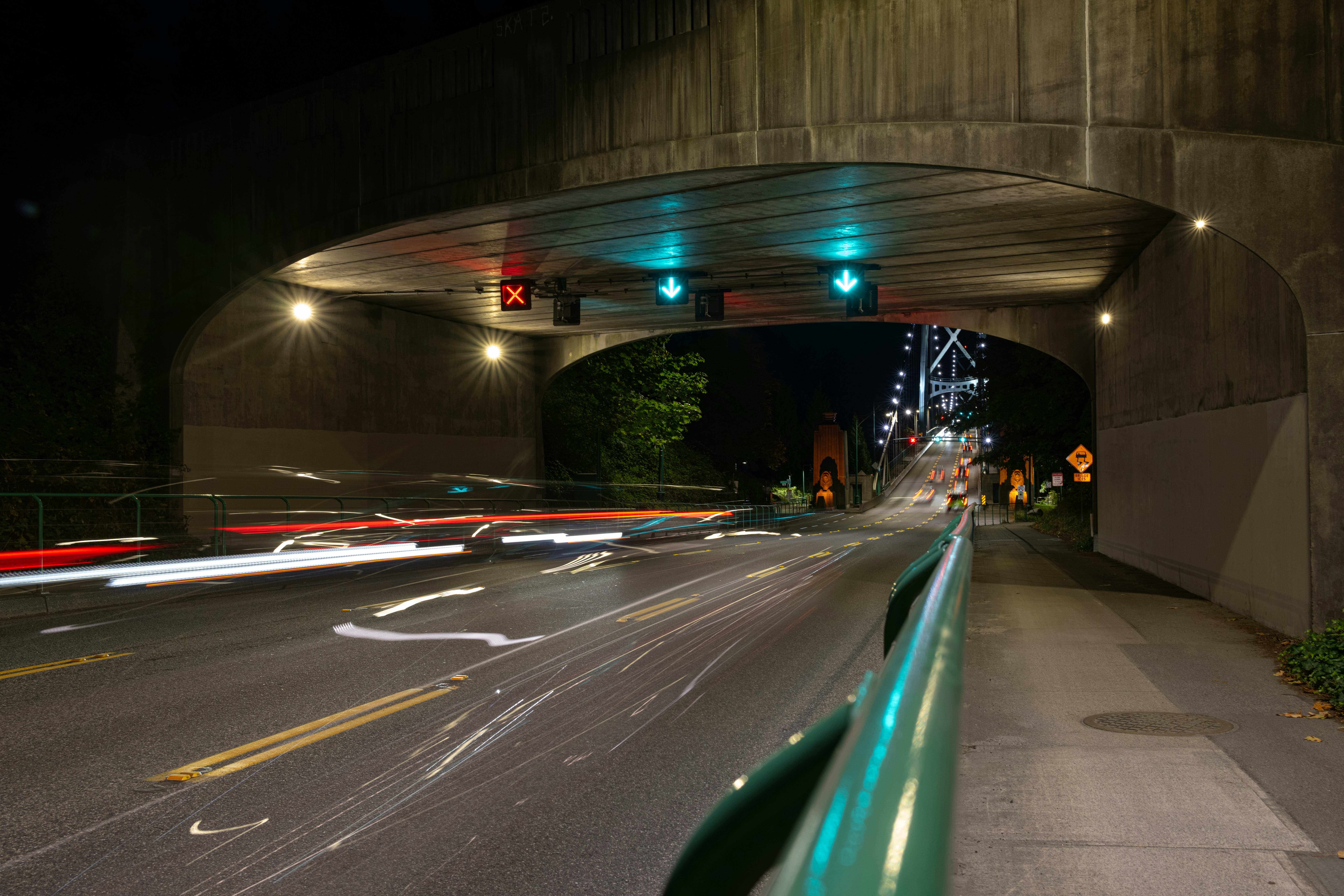 Long-exposure scene under a bridge shows light trails from moving traffic and a green railing along a nighttime street. A distant cityscape and illuminated signs peek through the underpass.