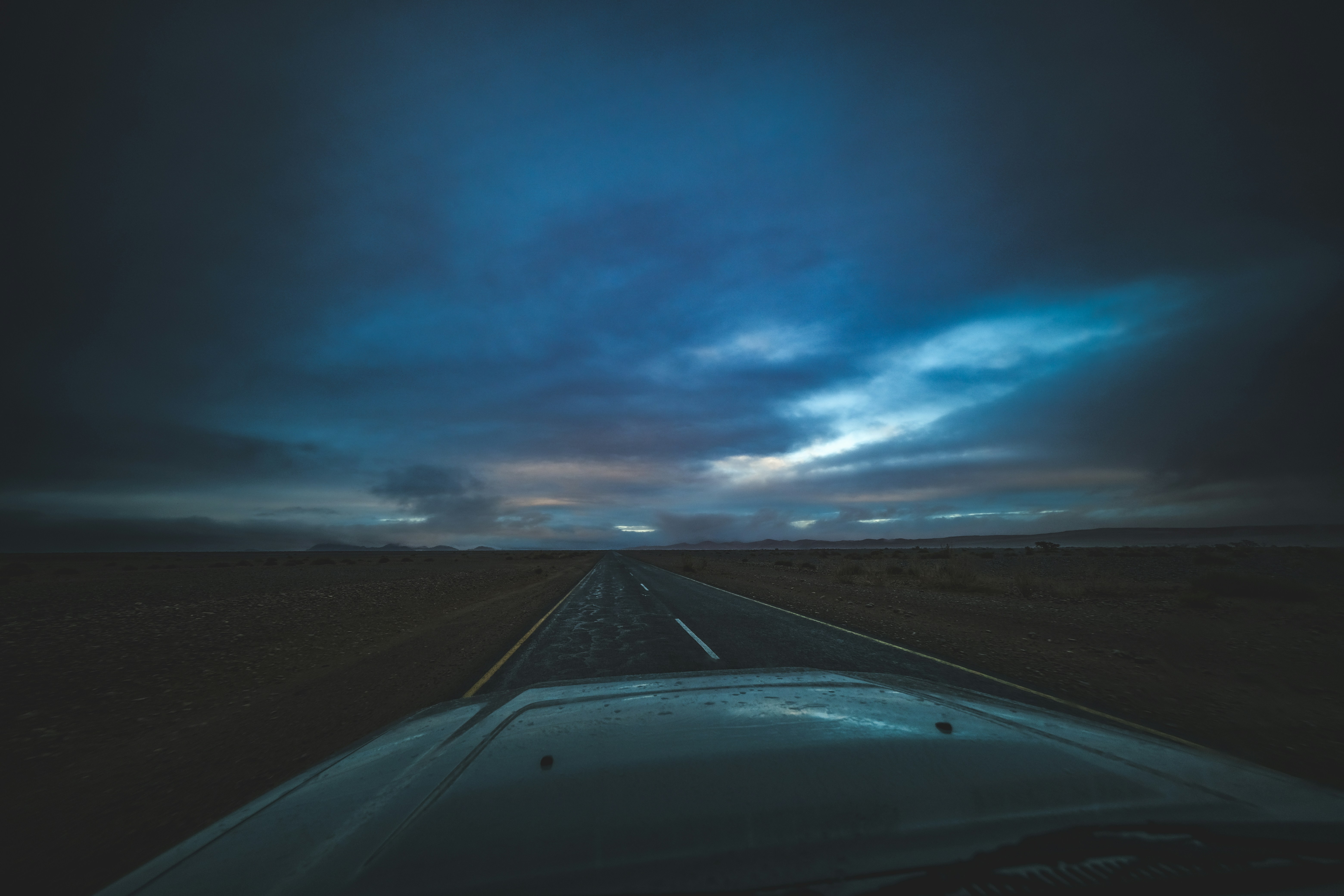 A view from a car hood onto a long road through a dark desert