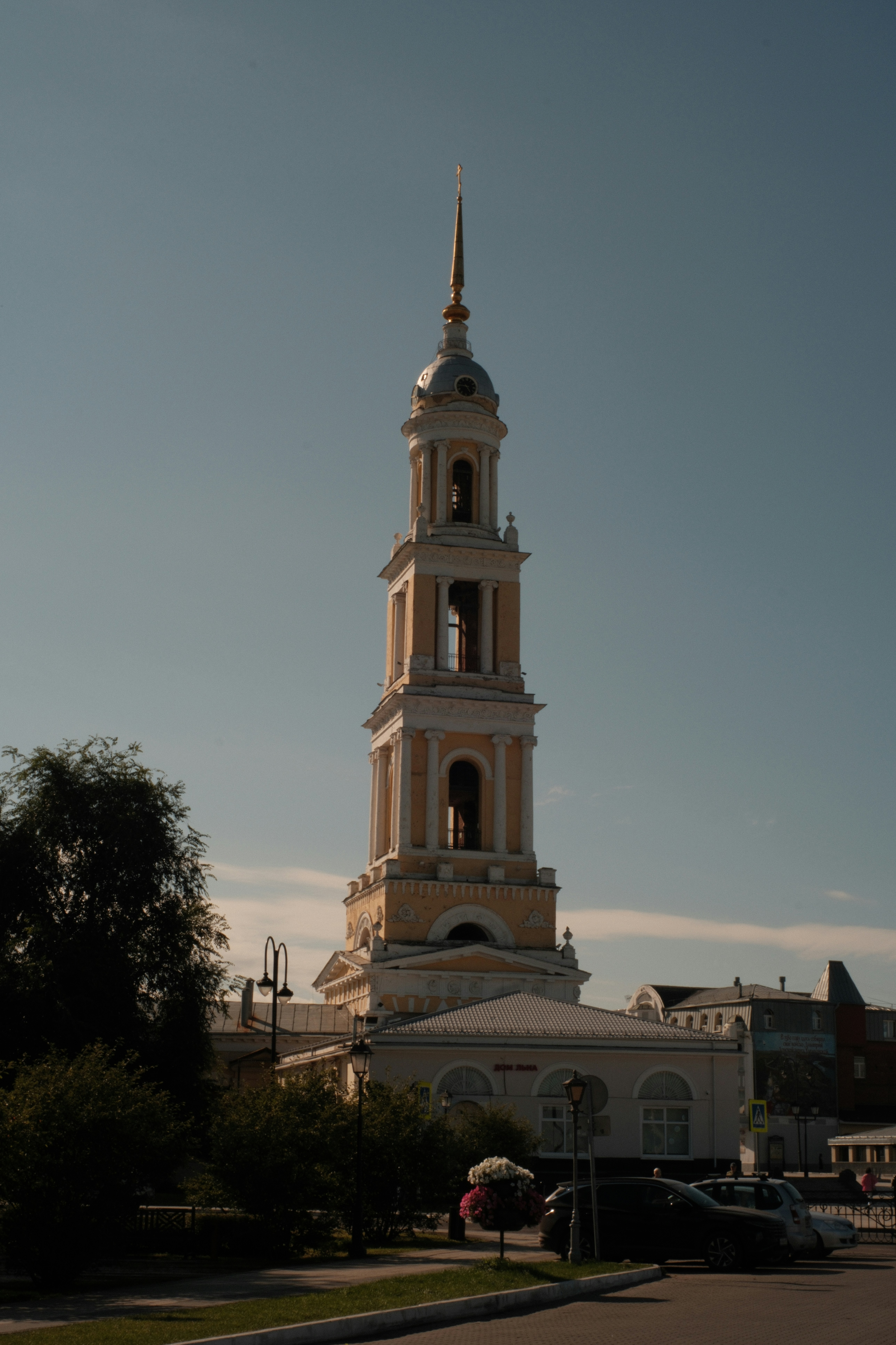 Sunlit bell tower rises above a low masonry complex; surrounding greenery and parked cars provide a quiet urban context. A single-structure architectural centerpiece stands against a clear blue sky.