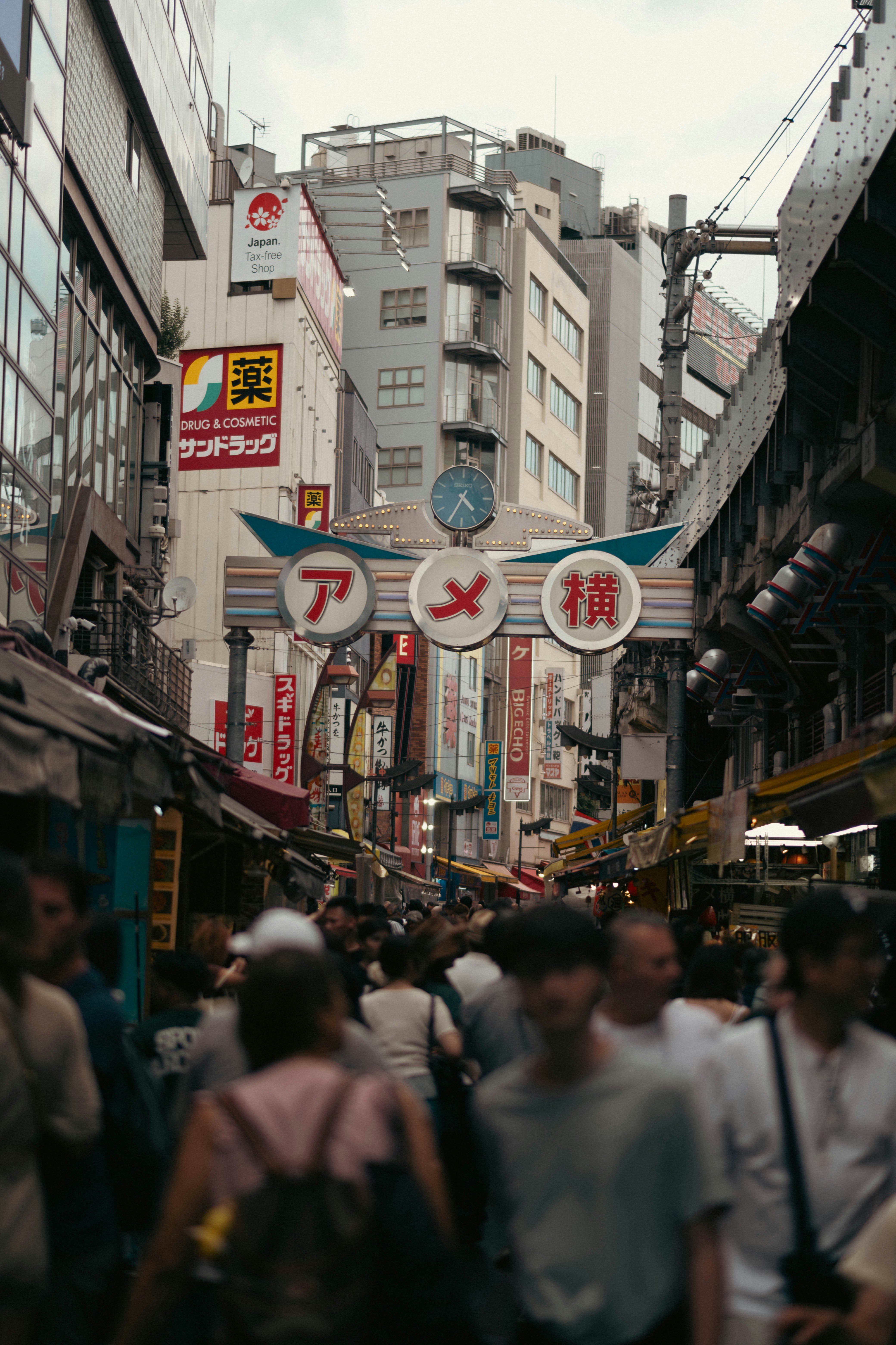 Bustling Ame-Yoko street scene with crowds beneath a retro sign arch in a narrow urban canyon; colorful storefronts frame a busy pedestrian corridor.