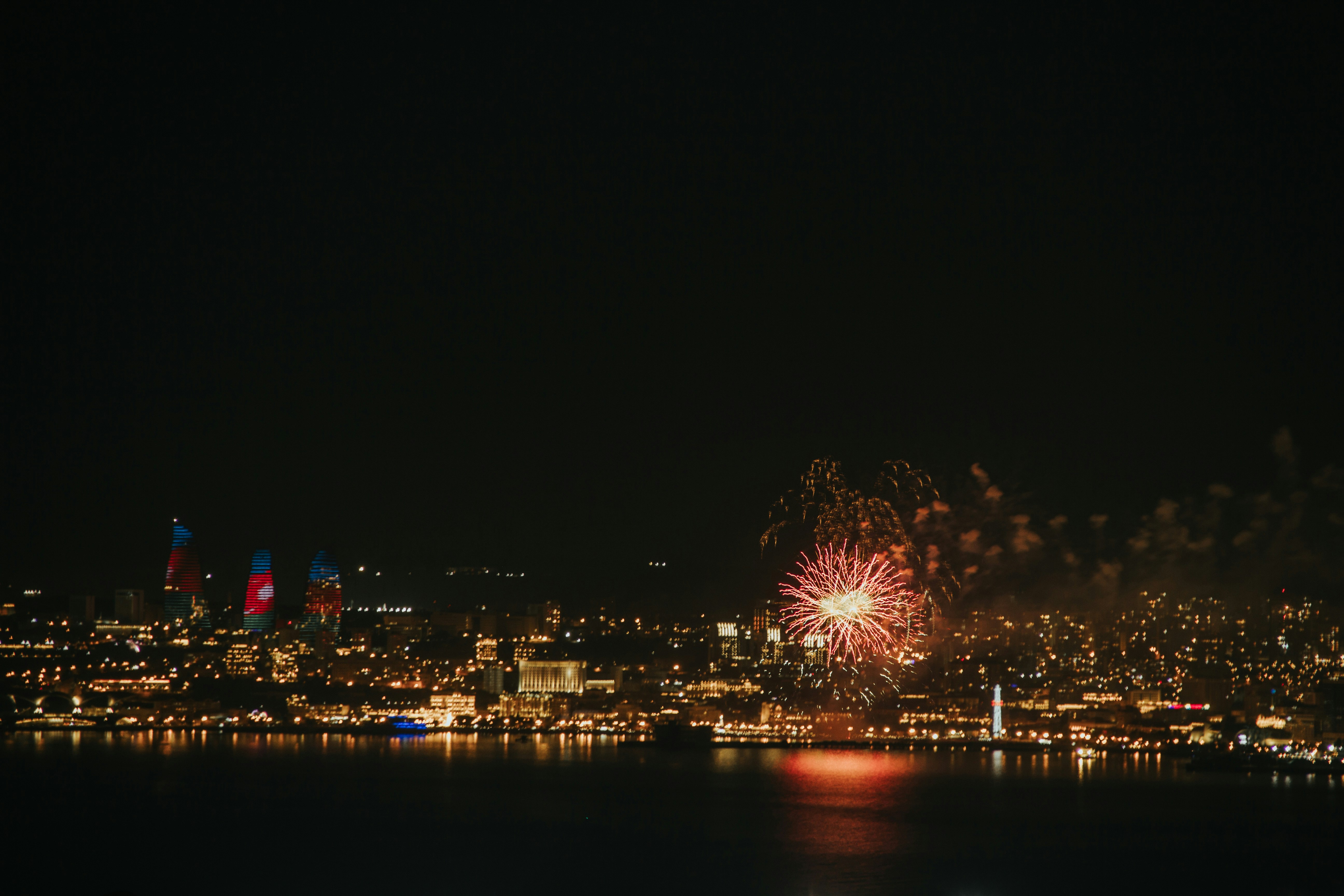 Dynamic night skyline with city lights and a prominent fireworks burst over the water, creating reflections across the harbor.