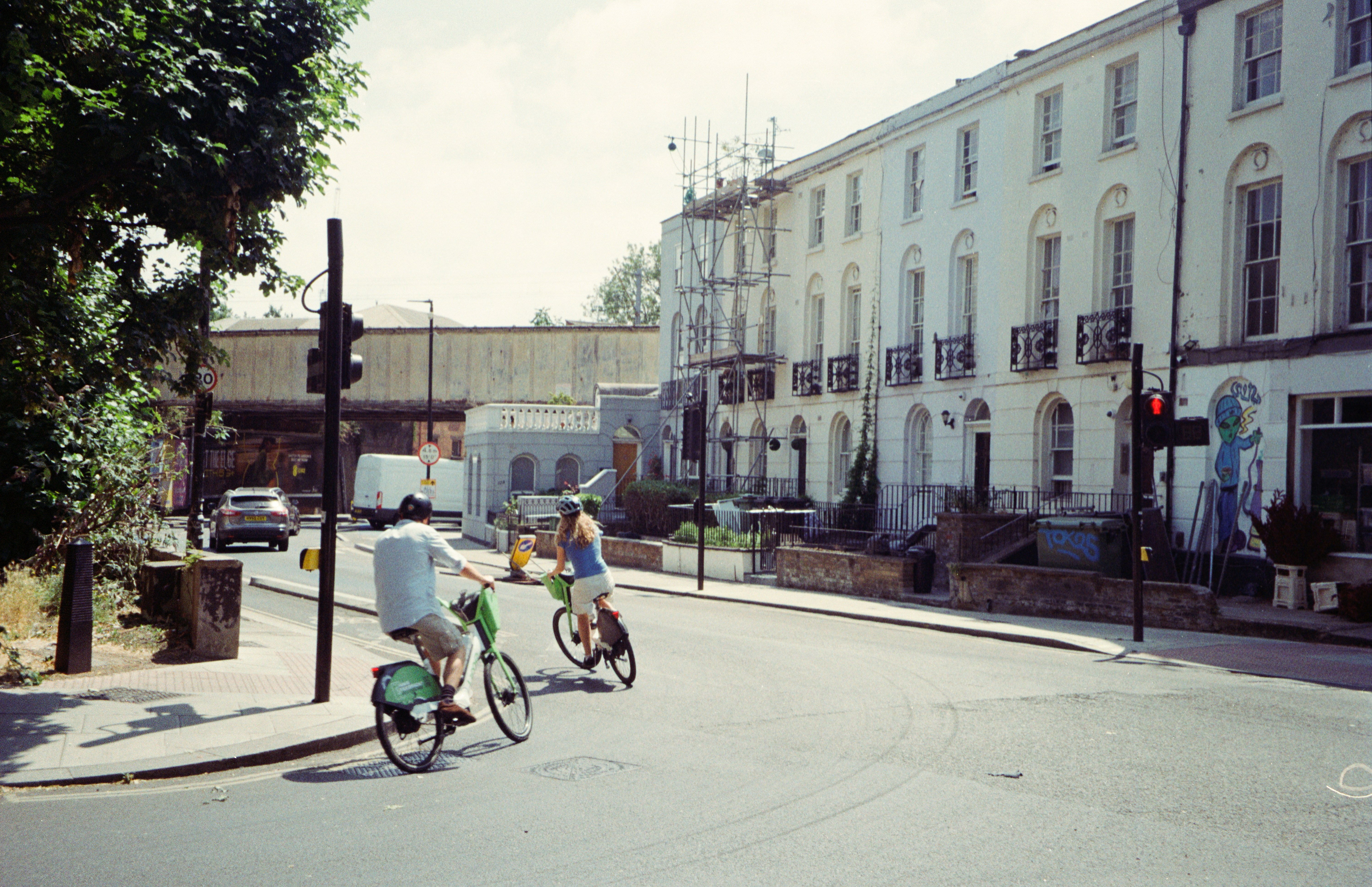 Two cyclists in a sunlit urban street bend past a row of pastel townhouses