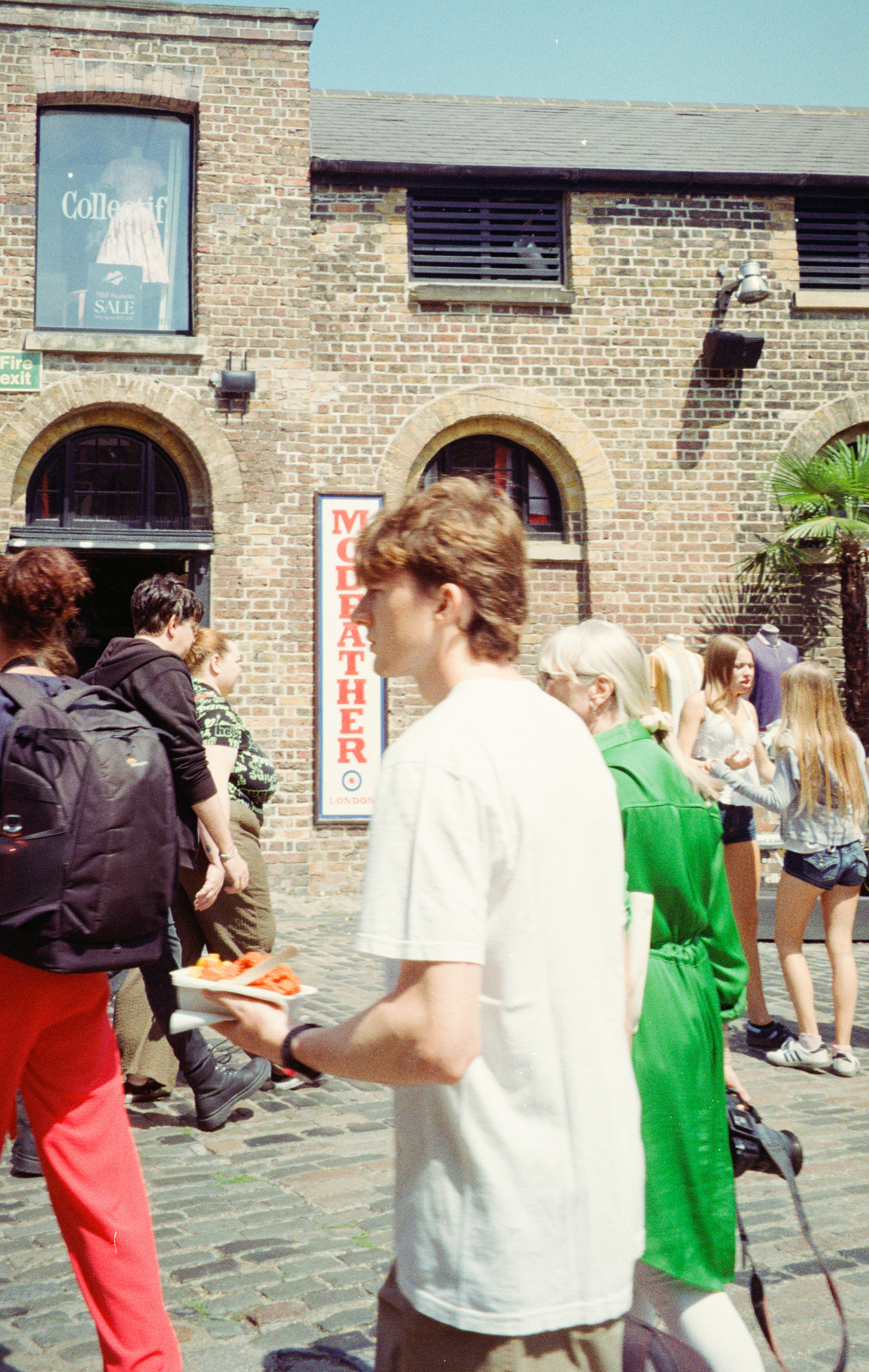 Crowd in a sunlit brick courtyard with a distracted photographer carrying snacks