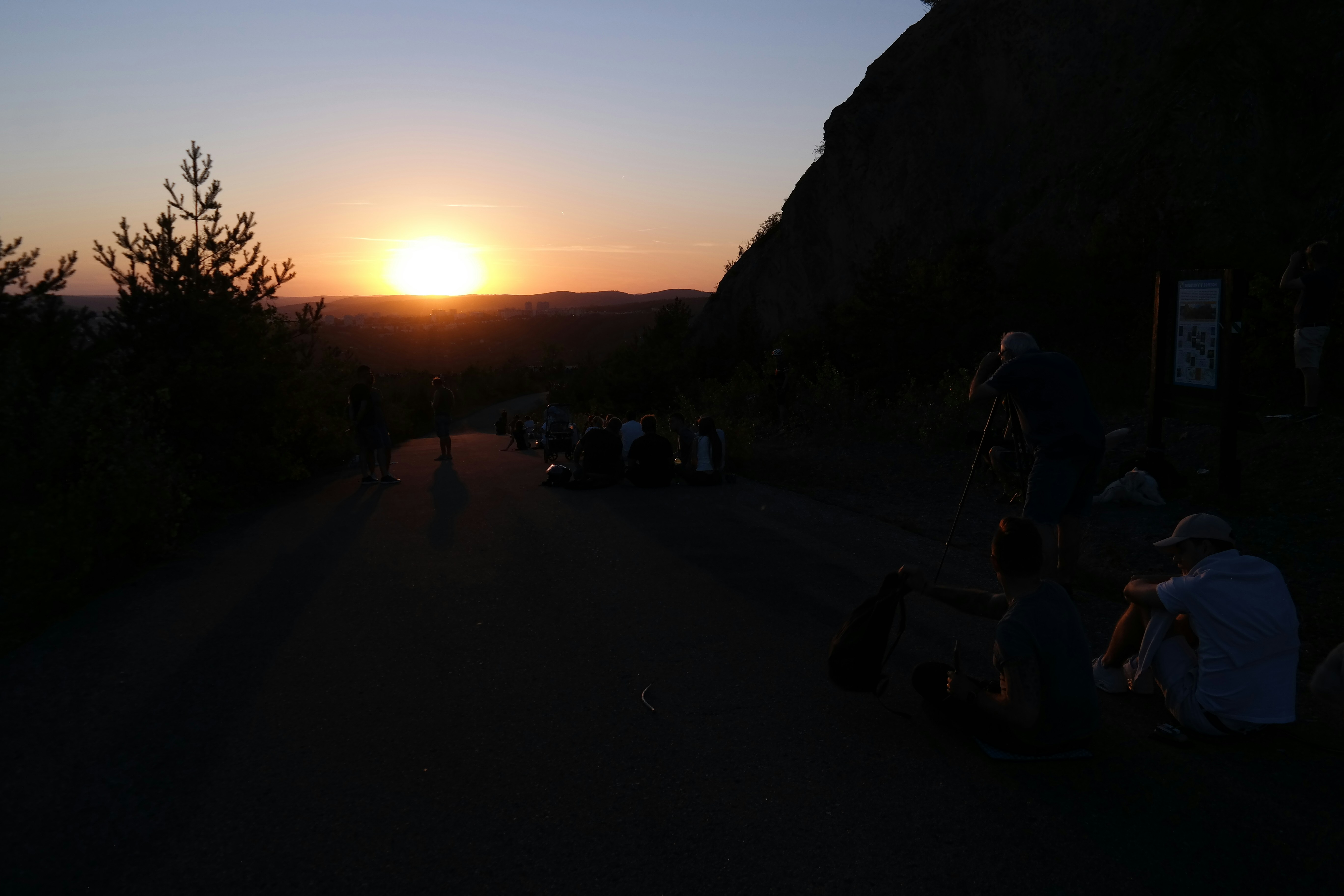 Horizon glow with silhouettes of people at a turnout, half-hidden by darkness; a rocky hillside on the right and trees on the left frame the scene.