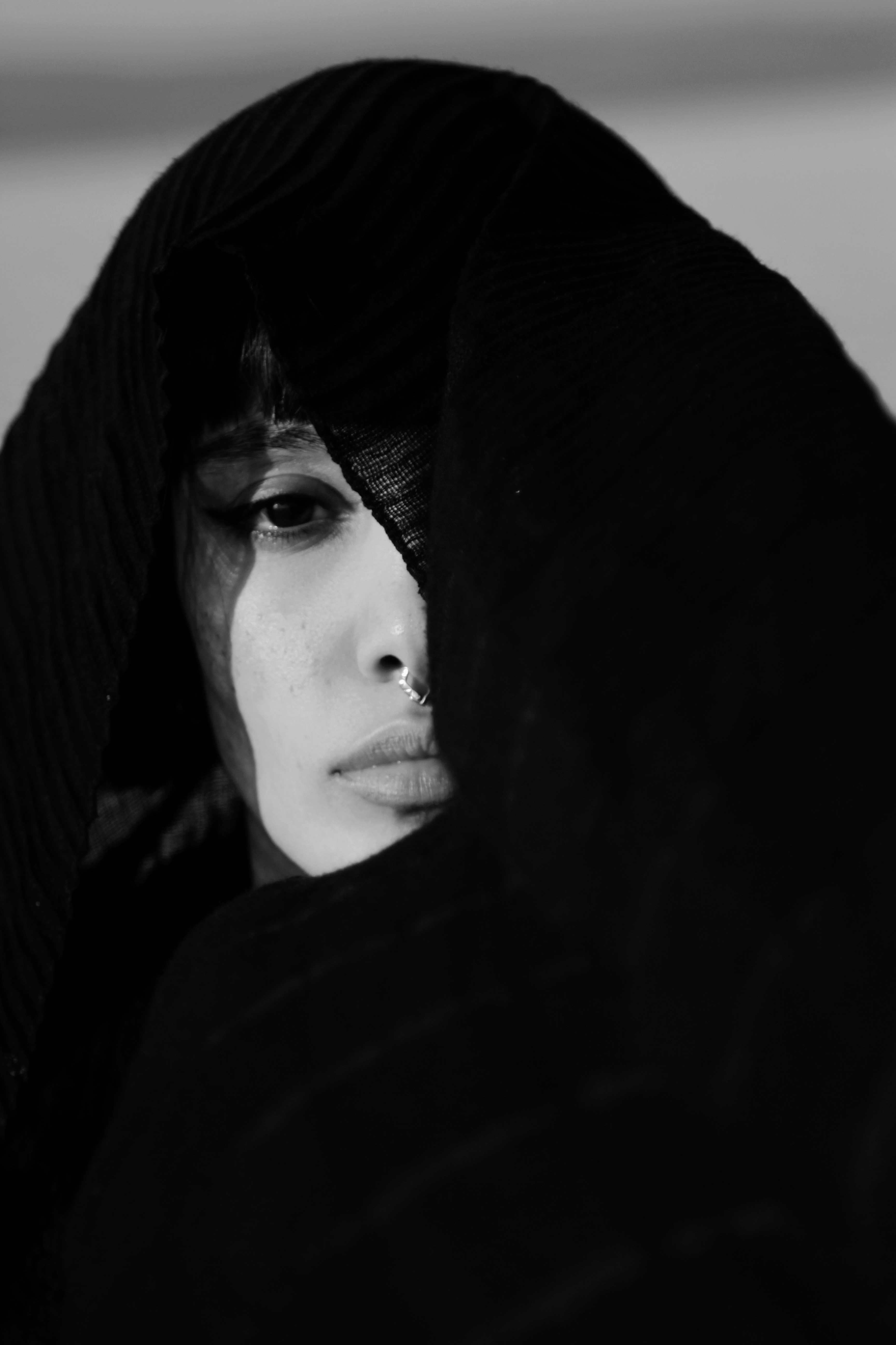 Close-up of a woman partially veiled in dark fabric, revealing a calm, piercing eye and subtle features; a minimalist, monochrome portrait emphasizing texture and mystery.