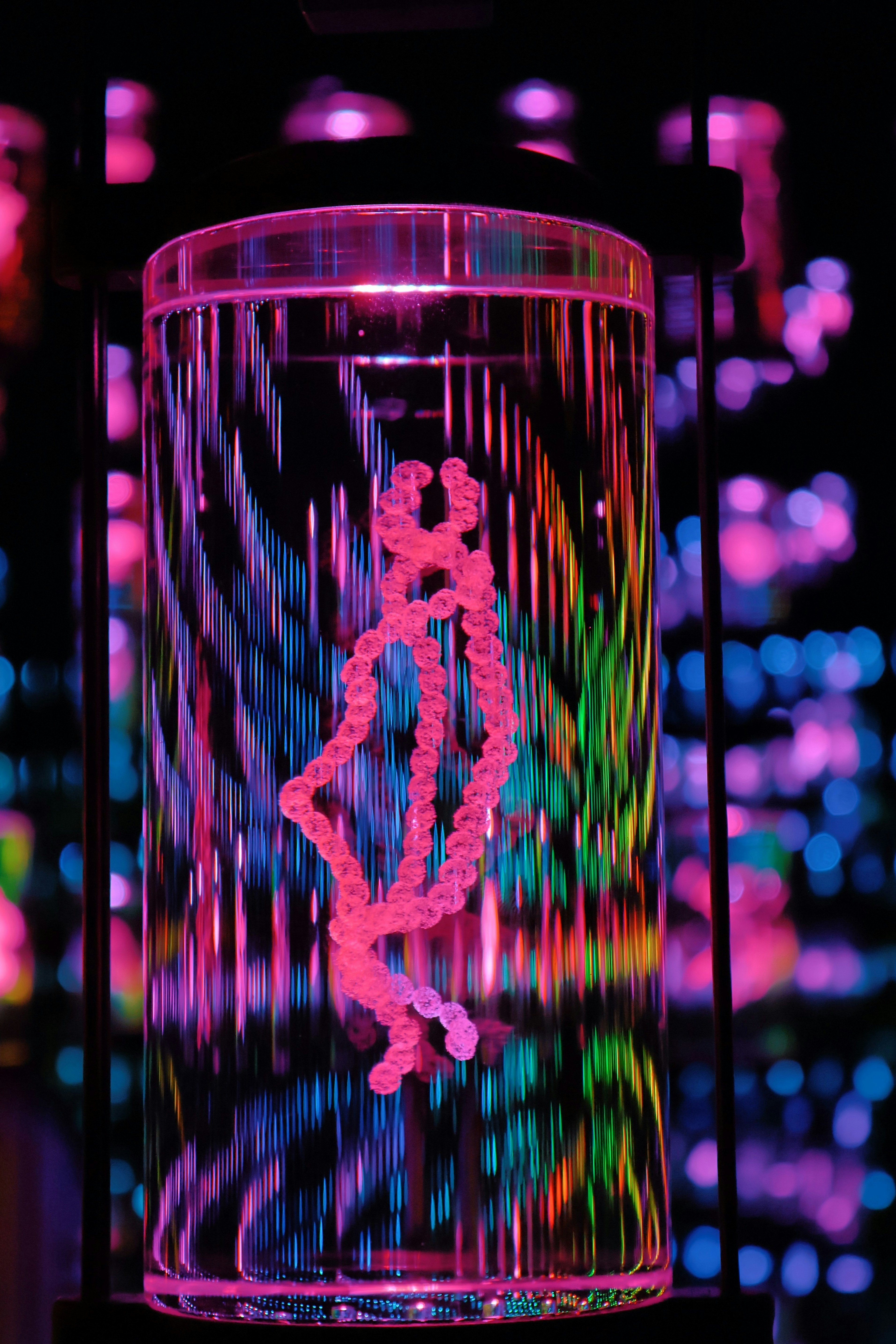 A tall cylindrical glass fixture glows with magenta and blue lights, revealing a pink beaded line drawing inside as its liquid-like surface distorts the vivid background. The scene is framed by dark stands, with a neon-bokeh backdrop suggesting a vibrant, club-like environment.