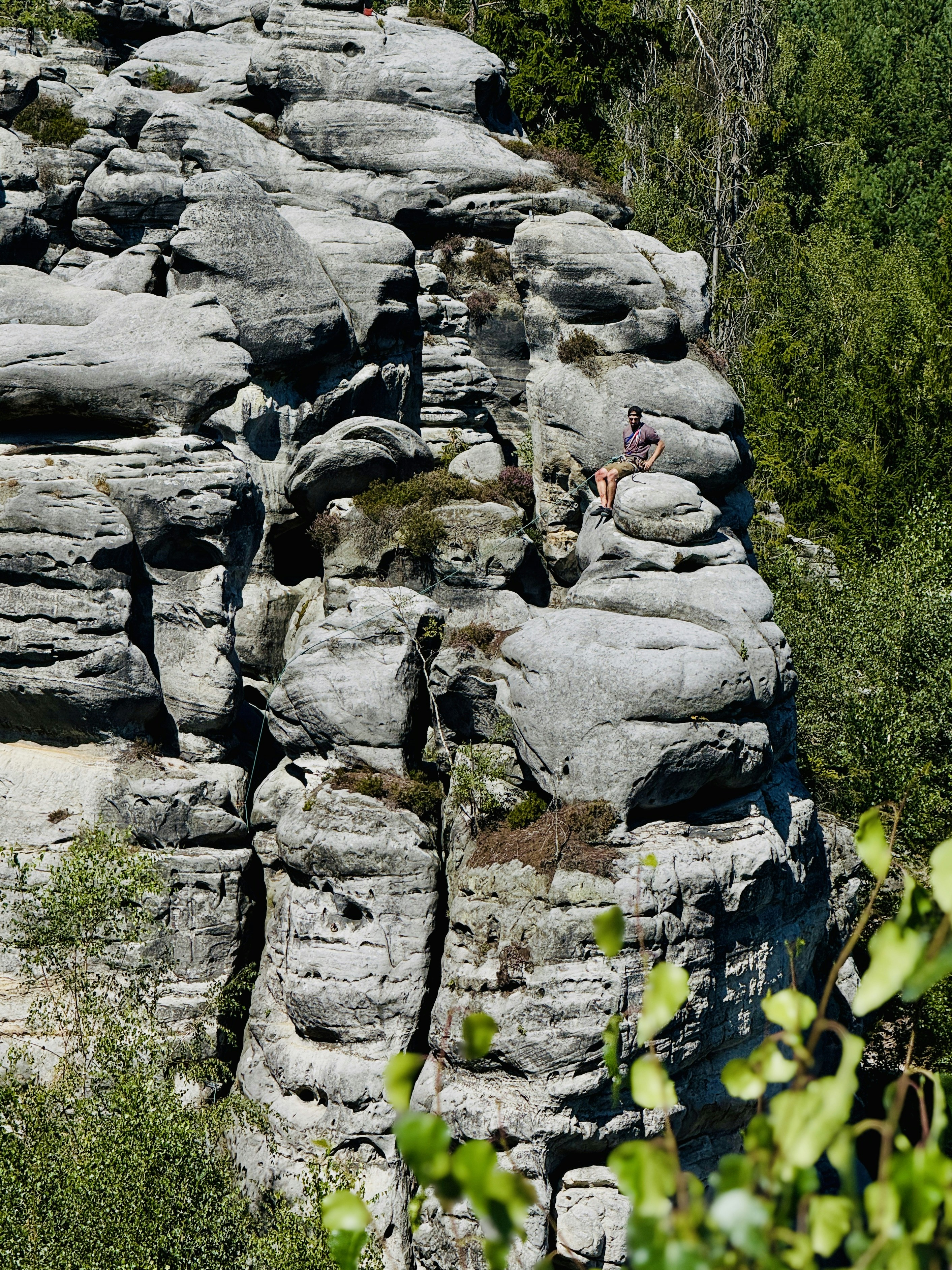 Rock spire with a lone climber seated on a ledge, surrounded by rugged granite and green forest. A quiet moment atop the formation as light highlights the textured cliff.