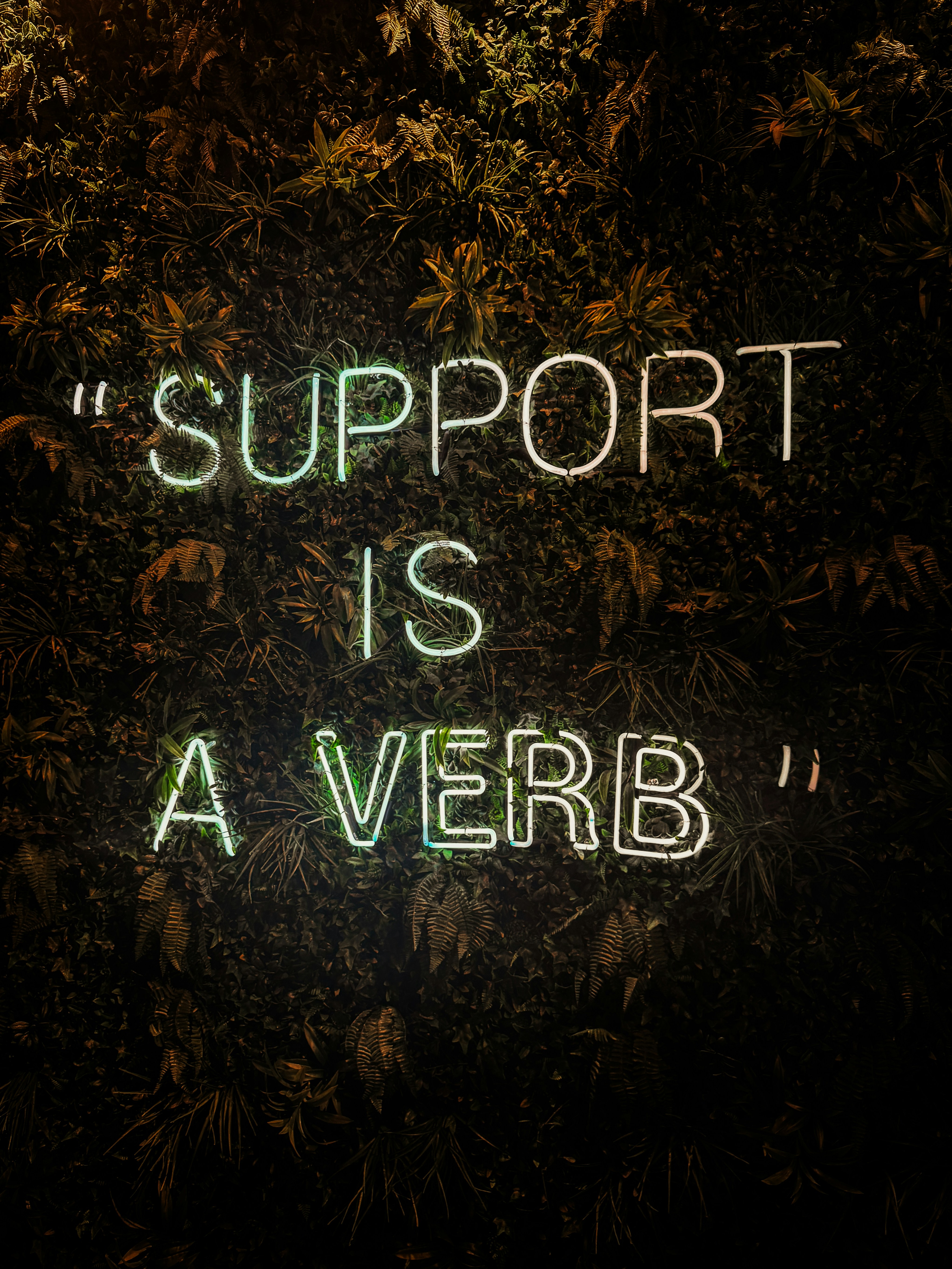 A neon sign reading "SUPPORT IS A VERB" on a dark green leafy wall