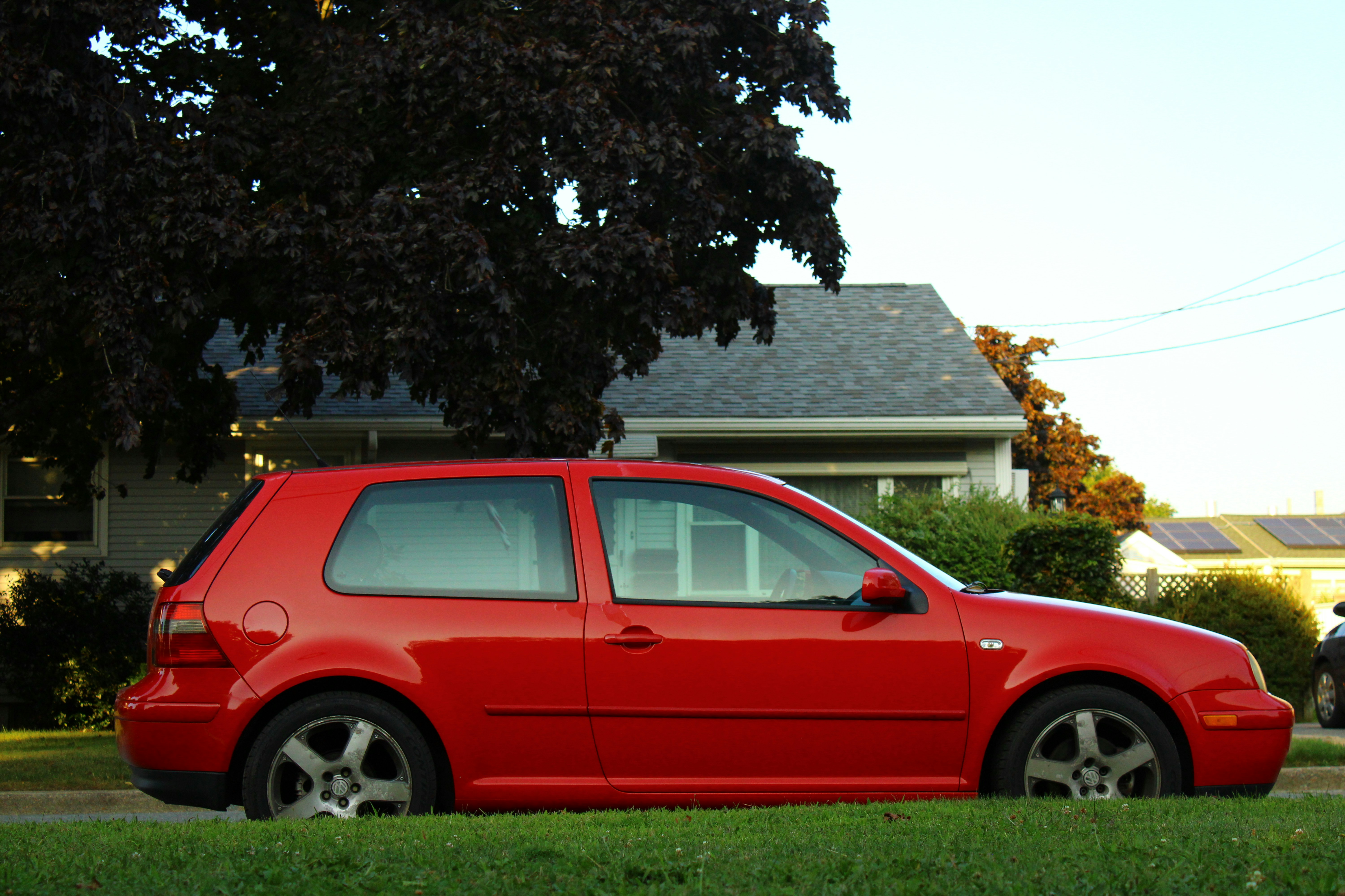 Red compact hatchback parked on a grassy verge beside a suburban house, with a large tree overhead and a clear sky backdrop.