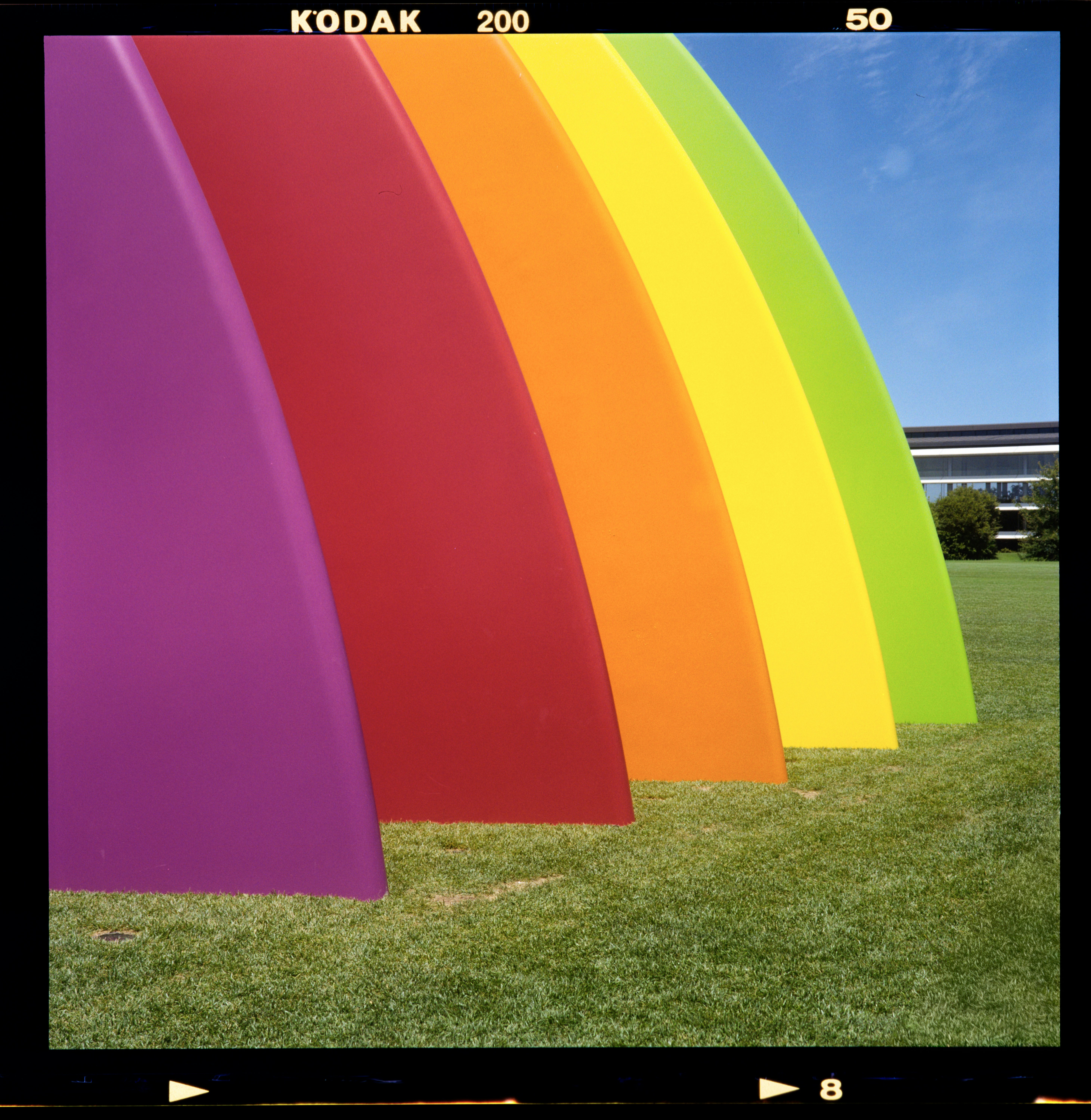 Bright rainbow arches stand along a grassy field under a blue sky; a modern outdoor sculpture with curved panels dominates the composition.