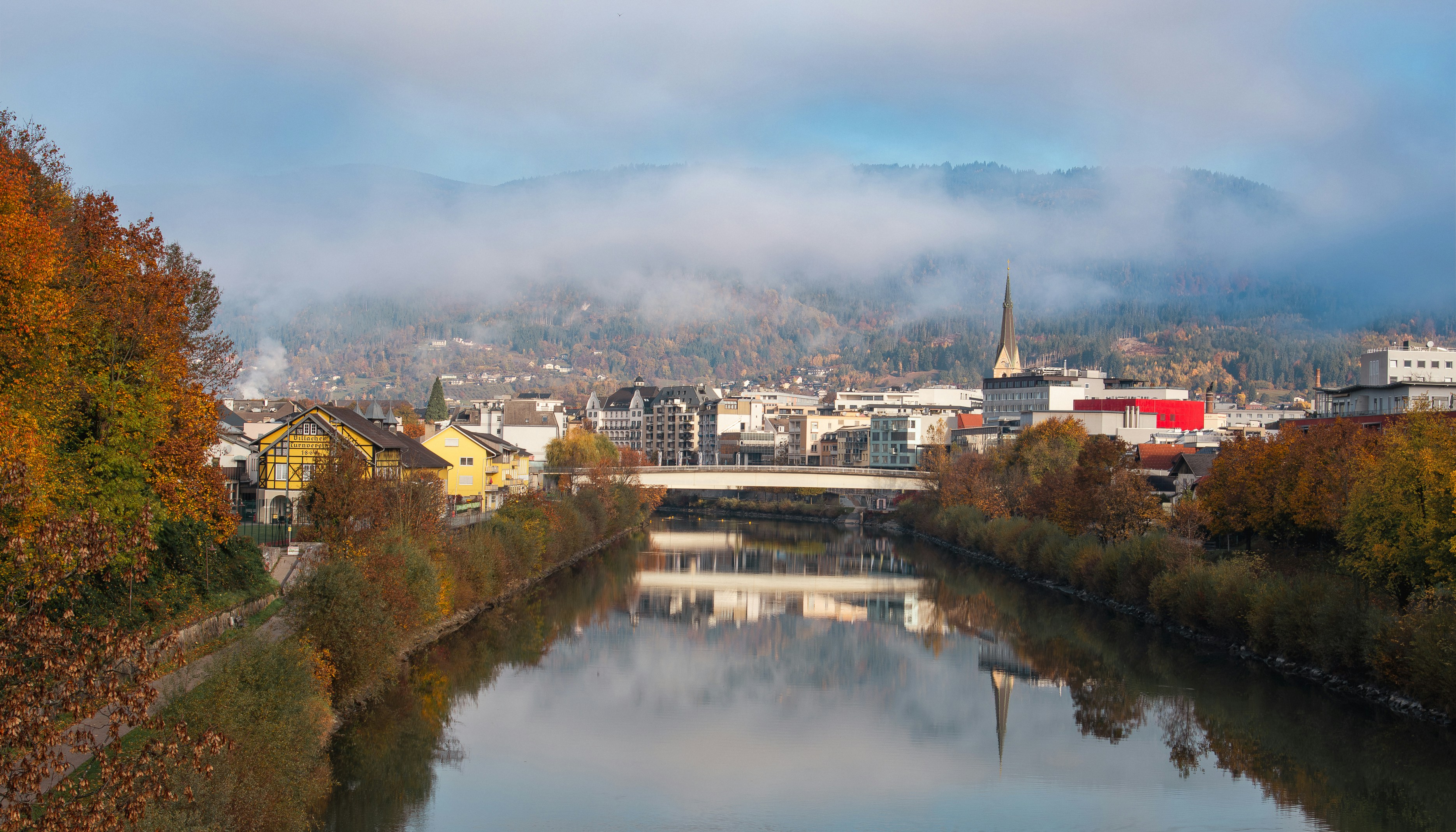 City river scene with autumnal trees, a bridge, and mist above the water reflecting the skyline. A tranquil urban landscape capturing seasonal color and calm waters.