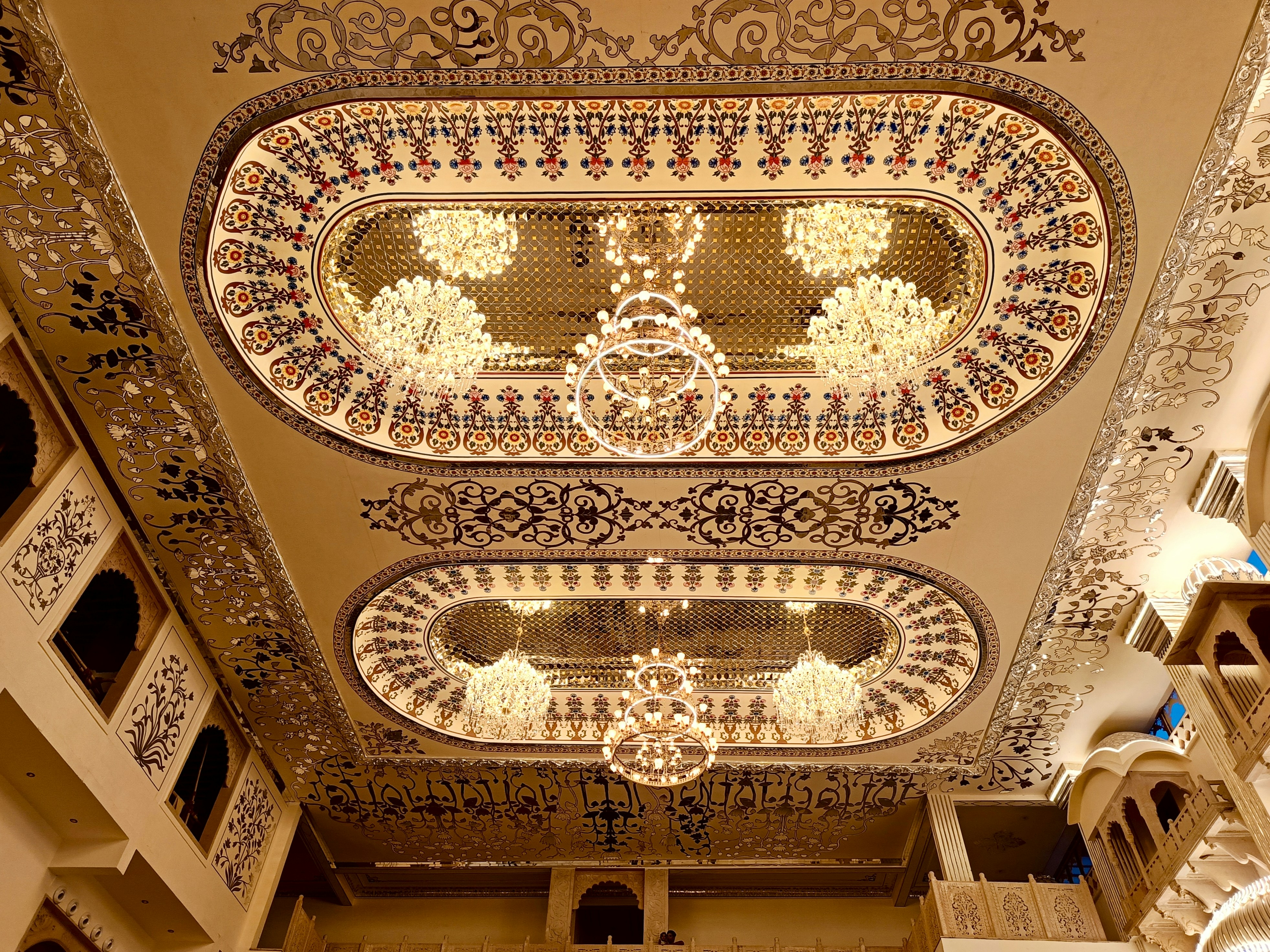 Ornate hotel ceiling with two inset domed panels, chandeliers, and intricate decorative patterns across warm-toned architecture.