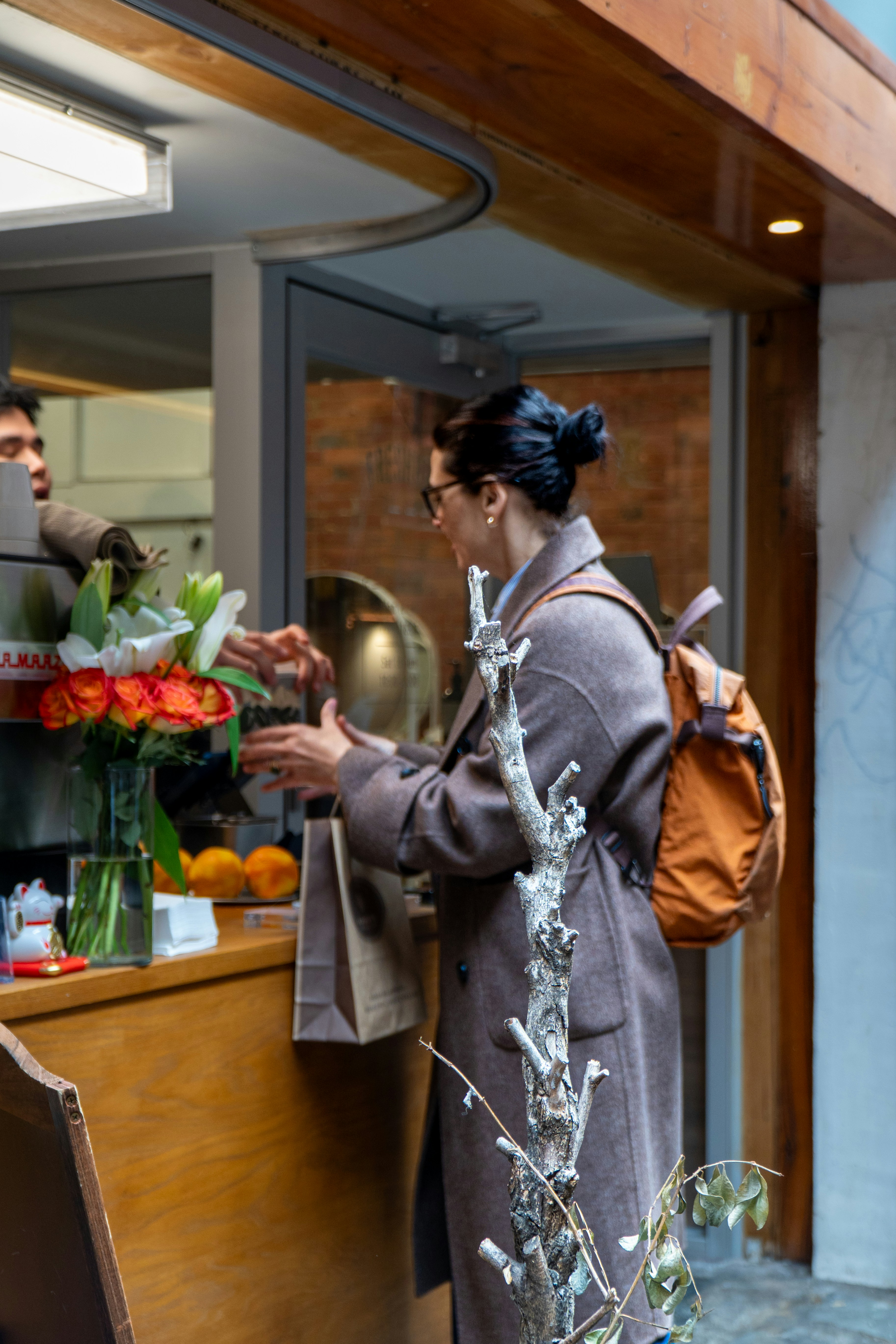 Woman in a gray coat with a backpack engages with a flower stall at a shop counter; a branch in foreground and warm interior lighting add depth.