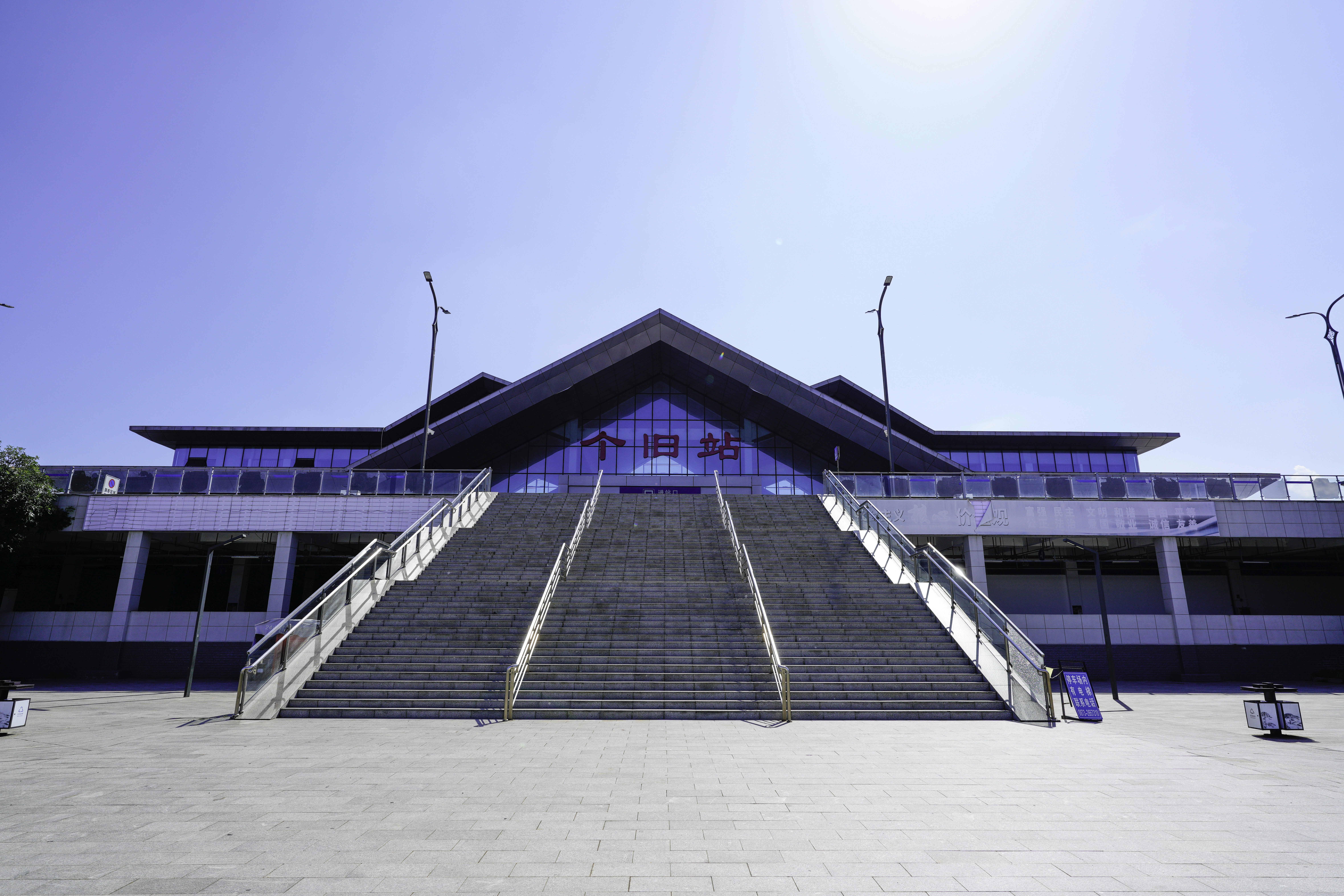 Wide-angle view of a symmetric station entrance with a central staircase leading to a peaked roof under a clear blue sky.