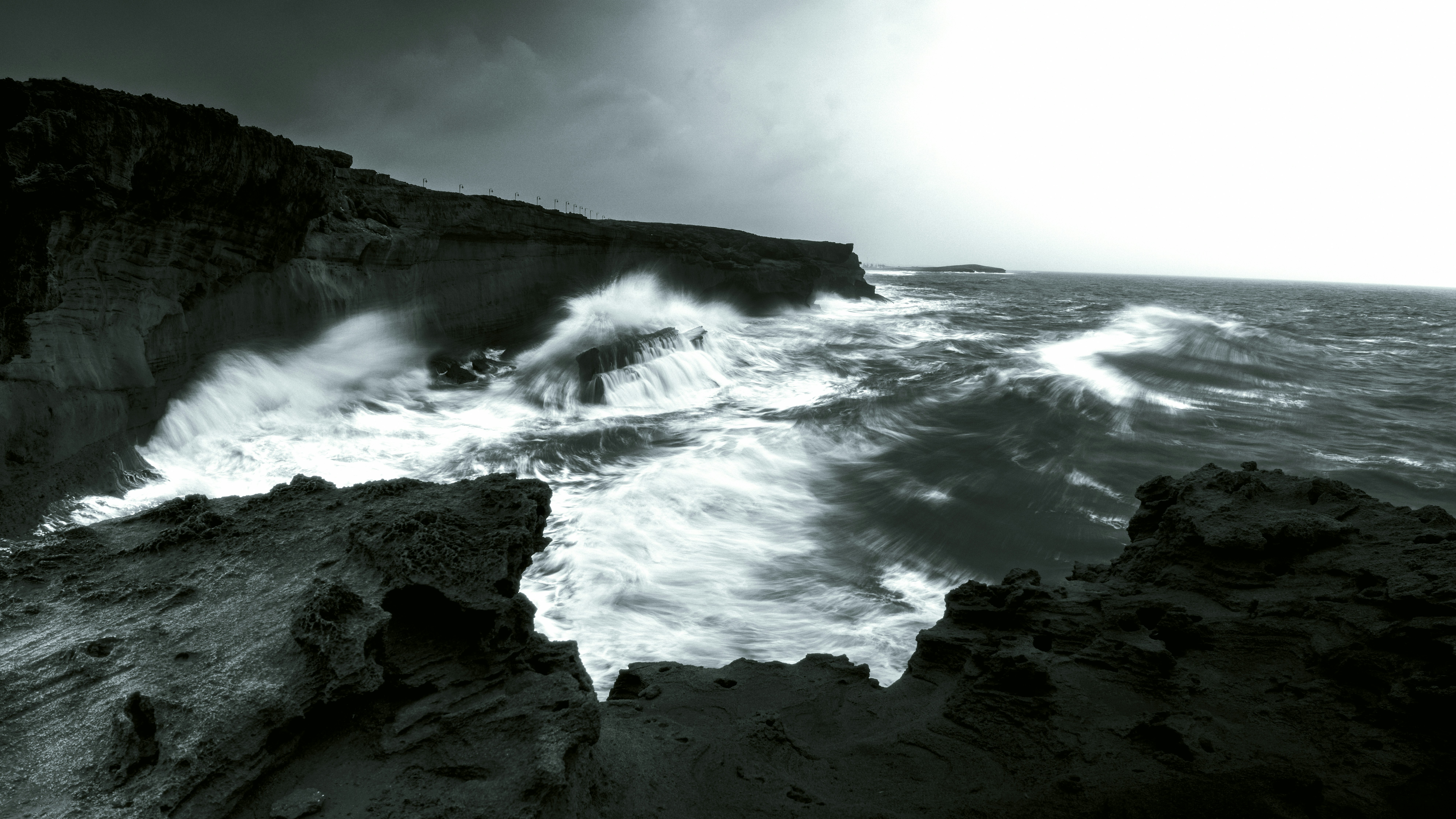 Dark coastal cliffs unleash white-tipped waves beneath a brooding sky, with turbulent sea traffic toward the open horizon.