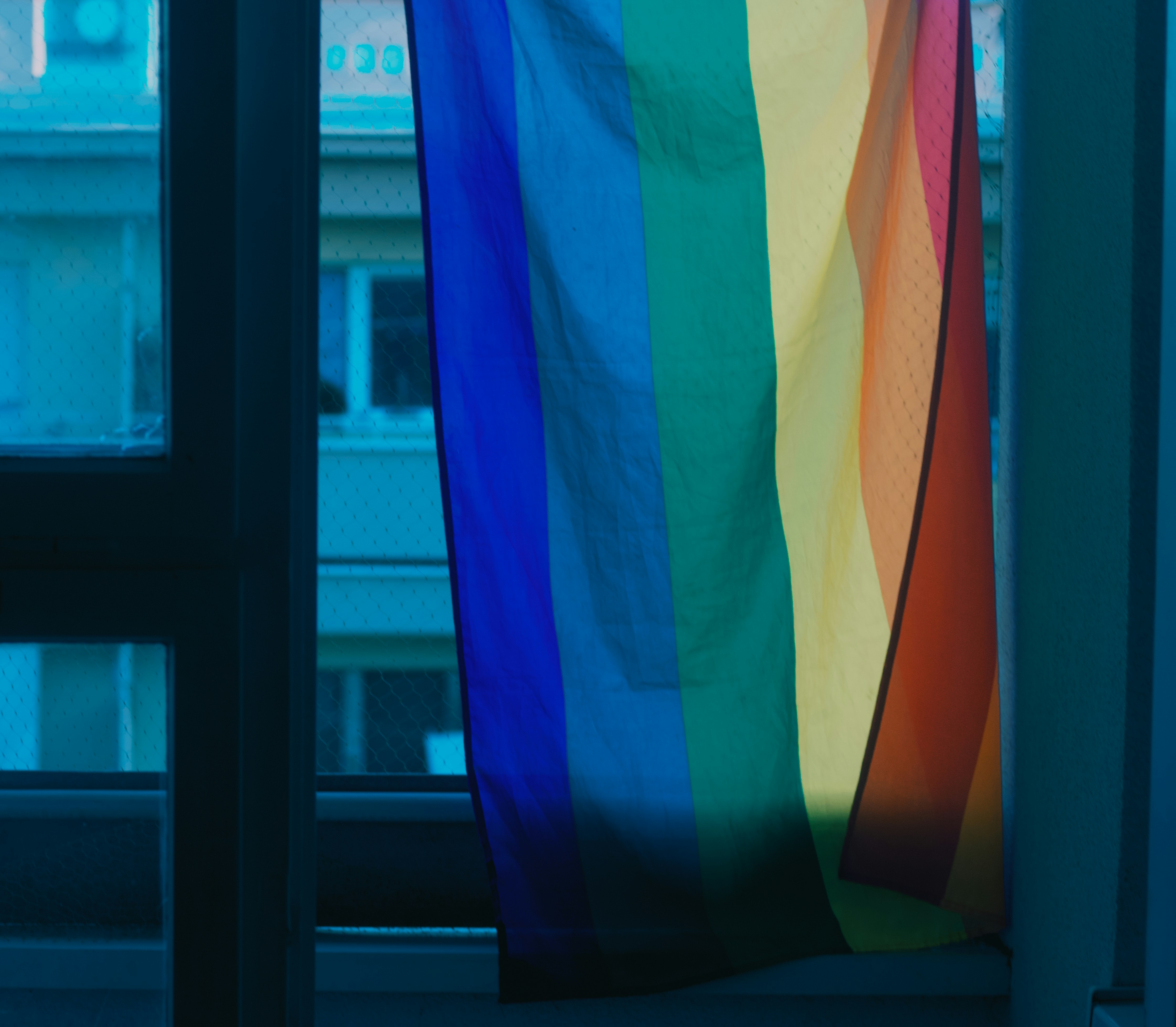 A rainbow flag drapes along a windowsill, its colors bold against the blue-tinted interior; the scene captures a quiet, colorful moment inside a room. This is a photographic capture of a fabric flag.