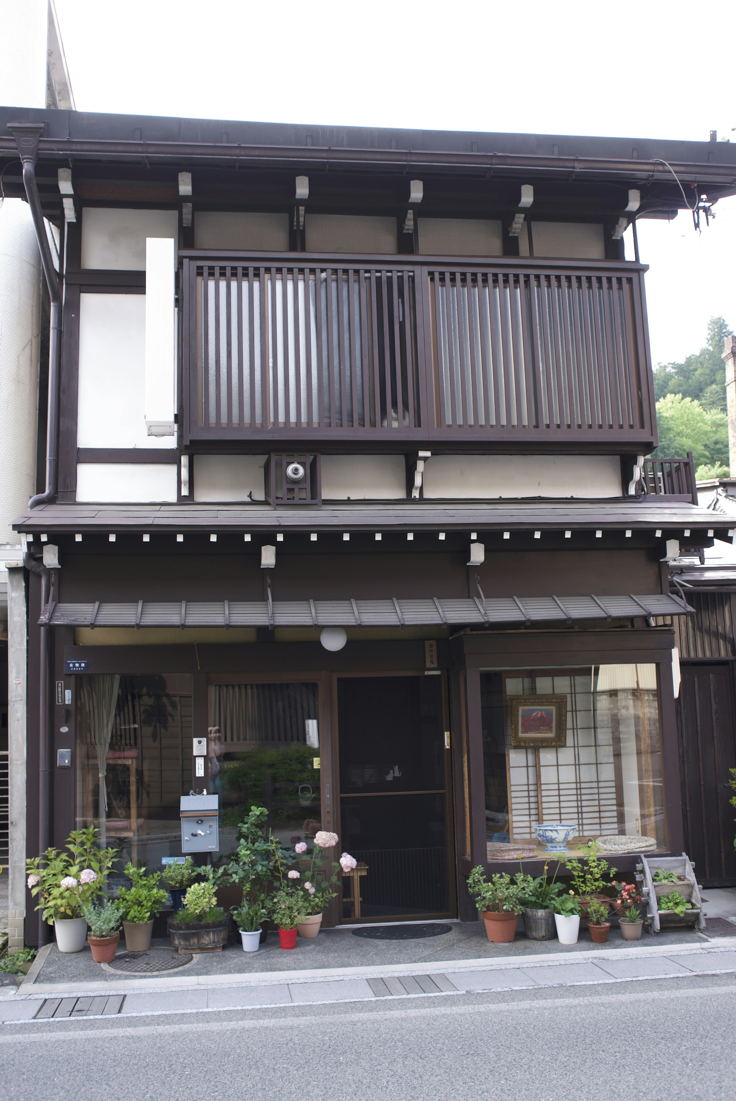 A two-story traditional Japanese shopfront with dark timber framing, sliding glass door, and an array of potted plants along the doorstep.