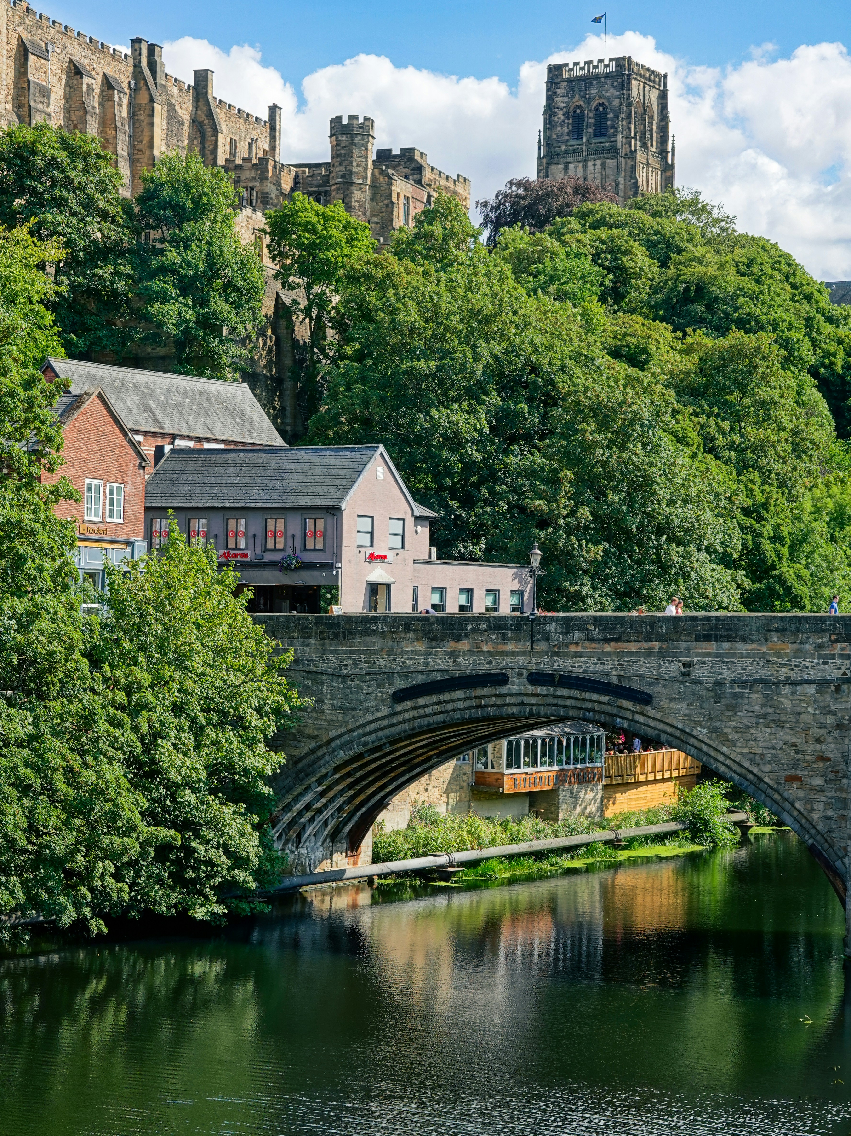 Stone bridge arches over a calm river with a cluster of brick buildings and lush trees behind, framed by a historic castle tower in the background. This is a photographic scene of urban-heritage landscape.