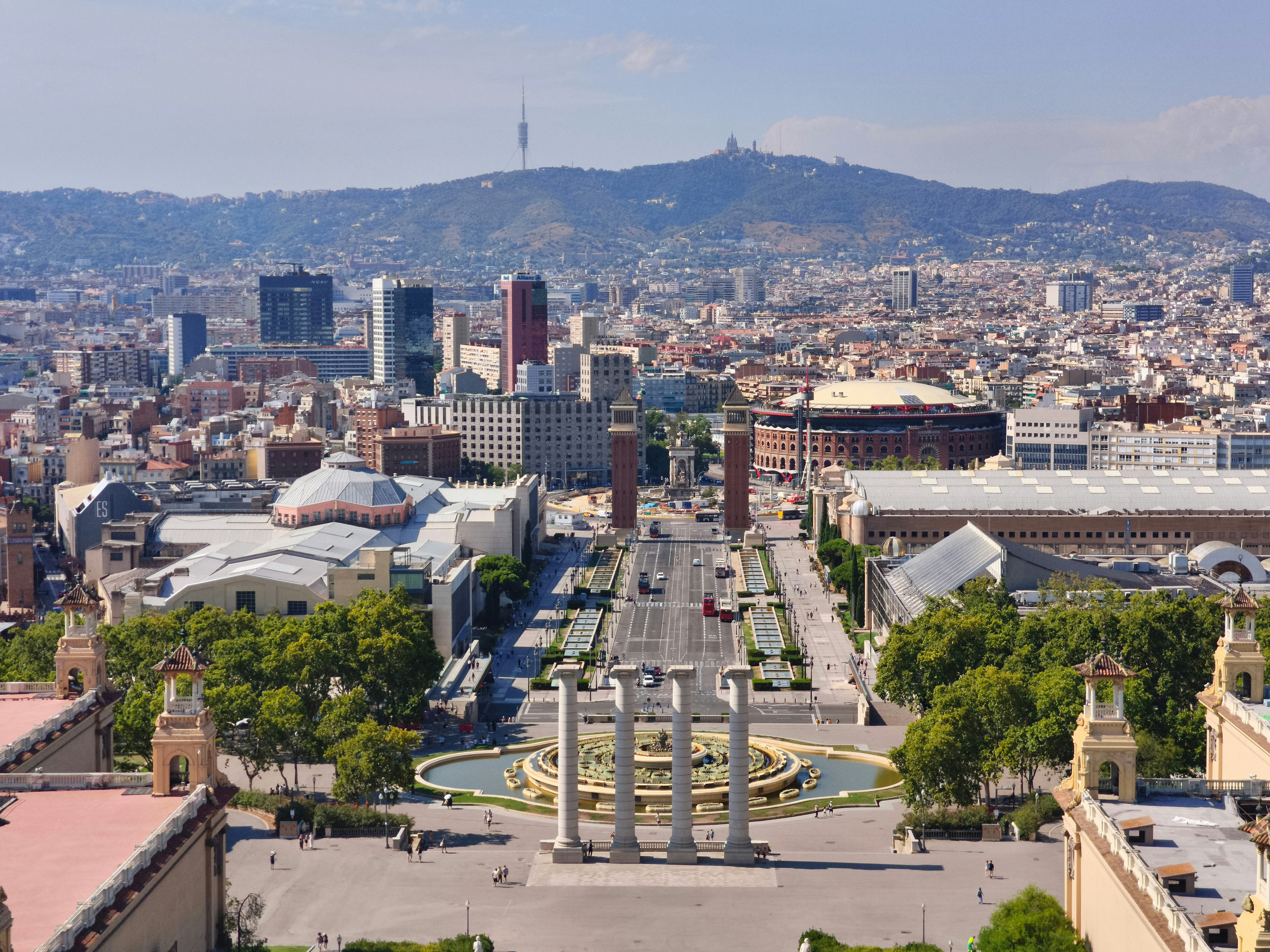 Wide-angle view of a grand city square with a central monument and running boulevard, flanked by stately buildings and green terraces, fading into distant hills.