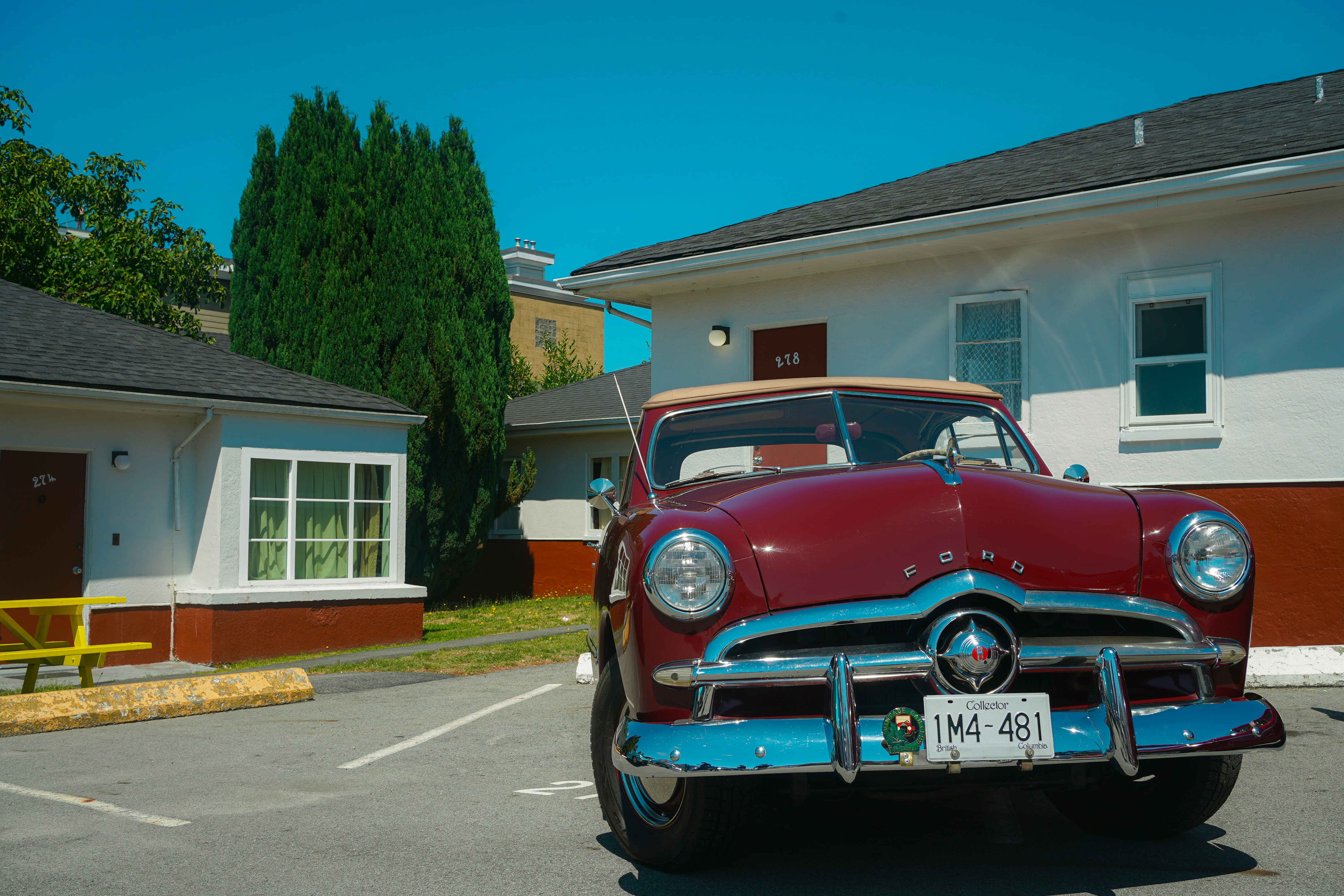A vintage red Ford sits bold in a sunlit driveway beside modest mid-century houses, chrome bumper gleaming and license plate visible. The scene conveys a nostalgic automotive moment in a calm suburban setting.