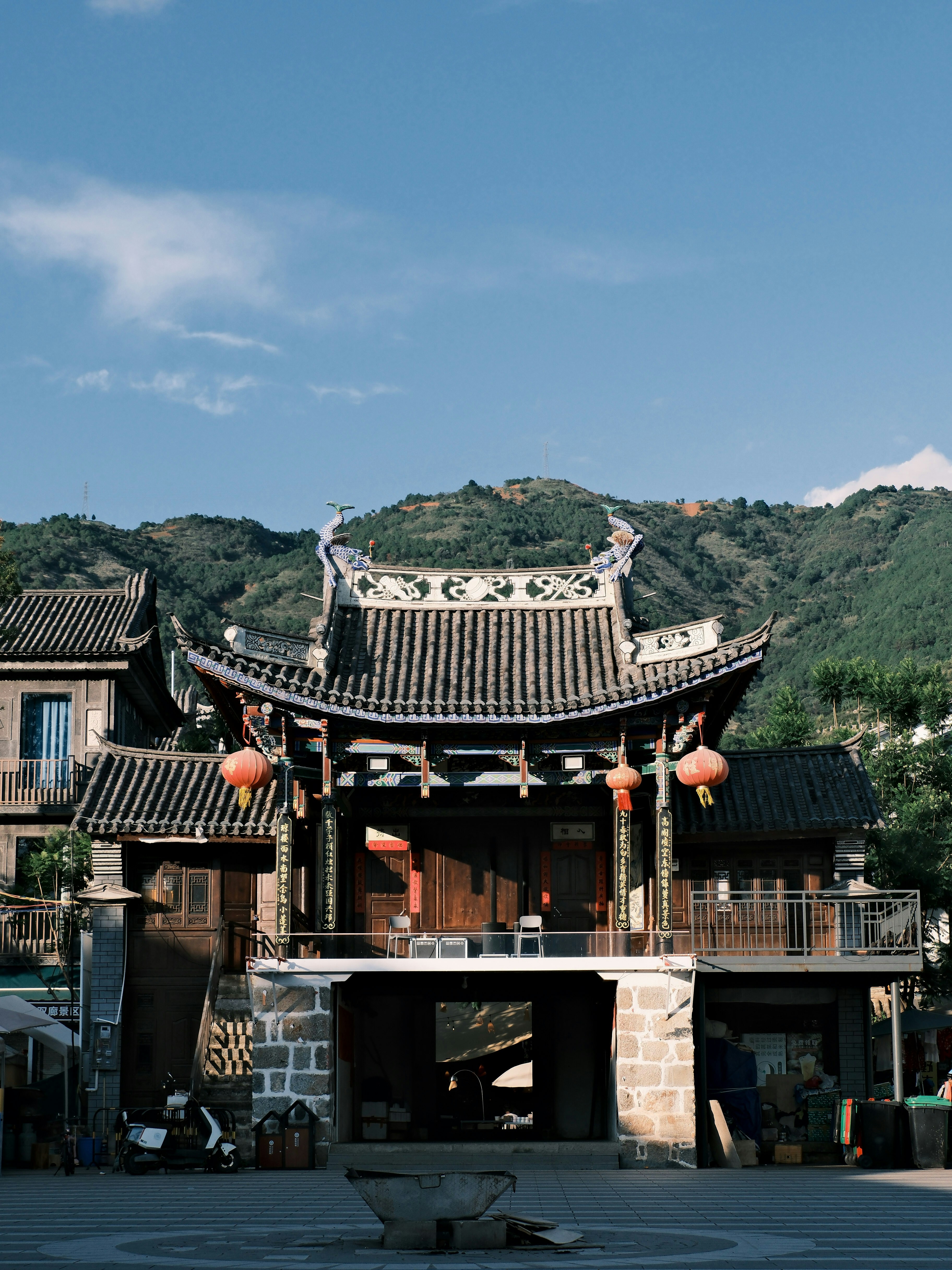 Traditional arched gateway with lanterns sits before a stone courtyard, set against rolling hills under a clear blue sky.