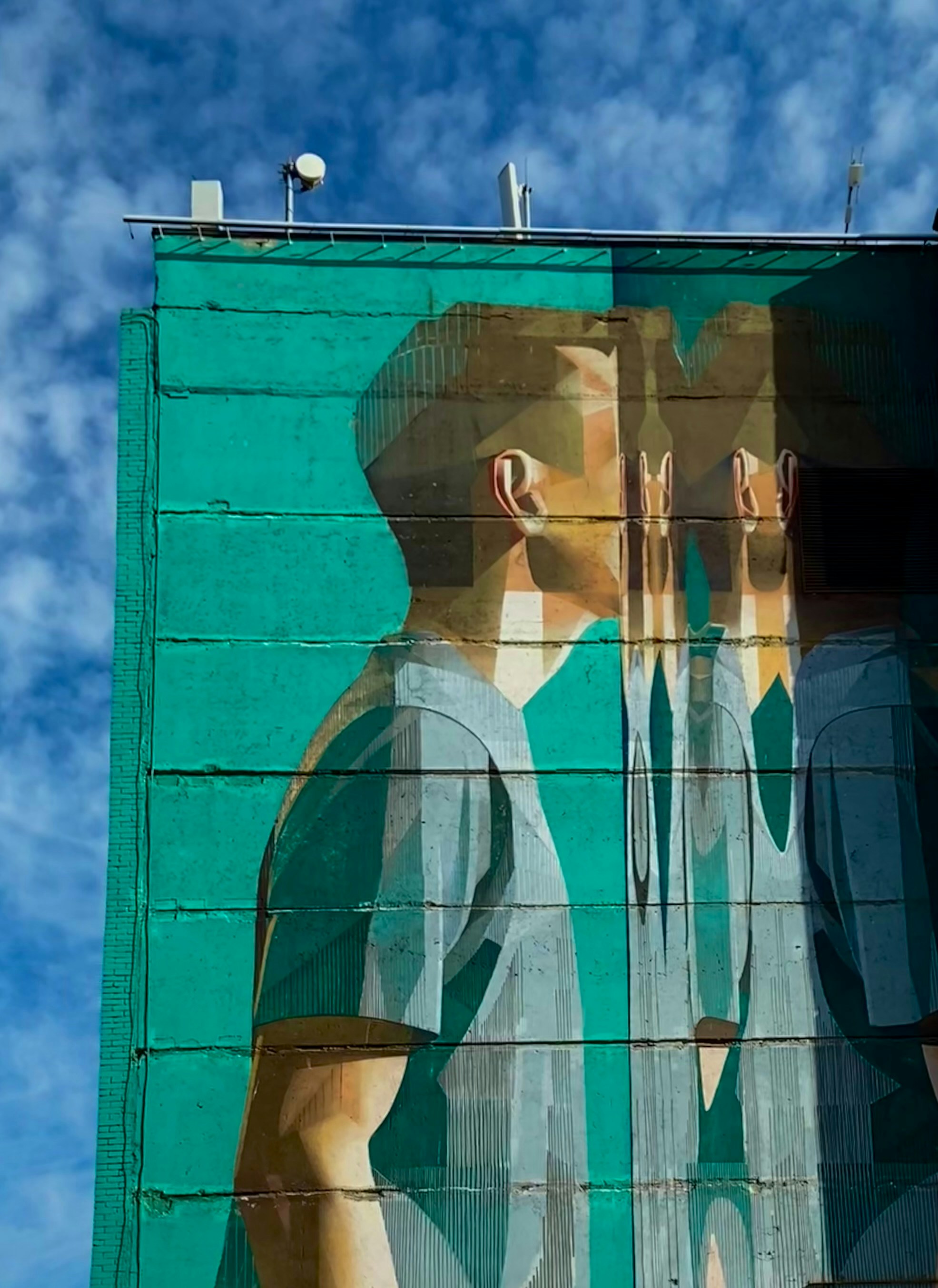The image features a large-scale geometric mural by Alexander Demkin on the side of an urban building. It depicts a stylized male figure in muted tones of teal, beige, and gray, rendered with angular facets and clean lines. A mirrored, fragmented silhouette overlays the central form, creating a sense of duality and reflection. The composition contrasts the sharp geometry of the artwork with the building’s industrial texture and the soft blue sky with wispy clouds.