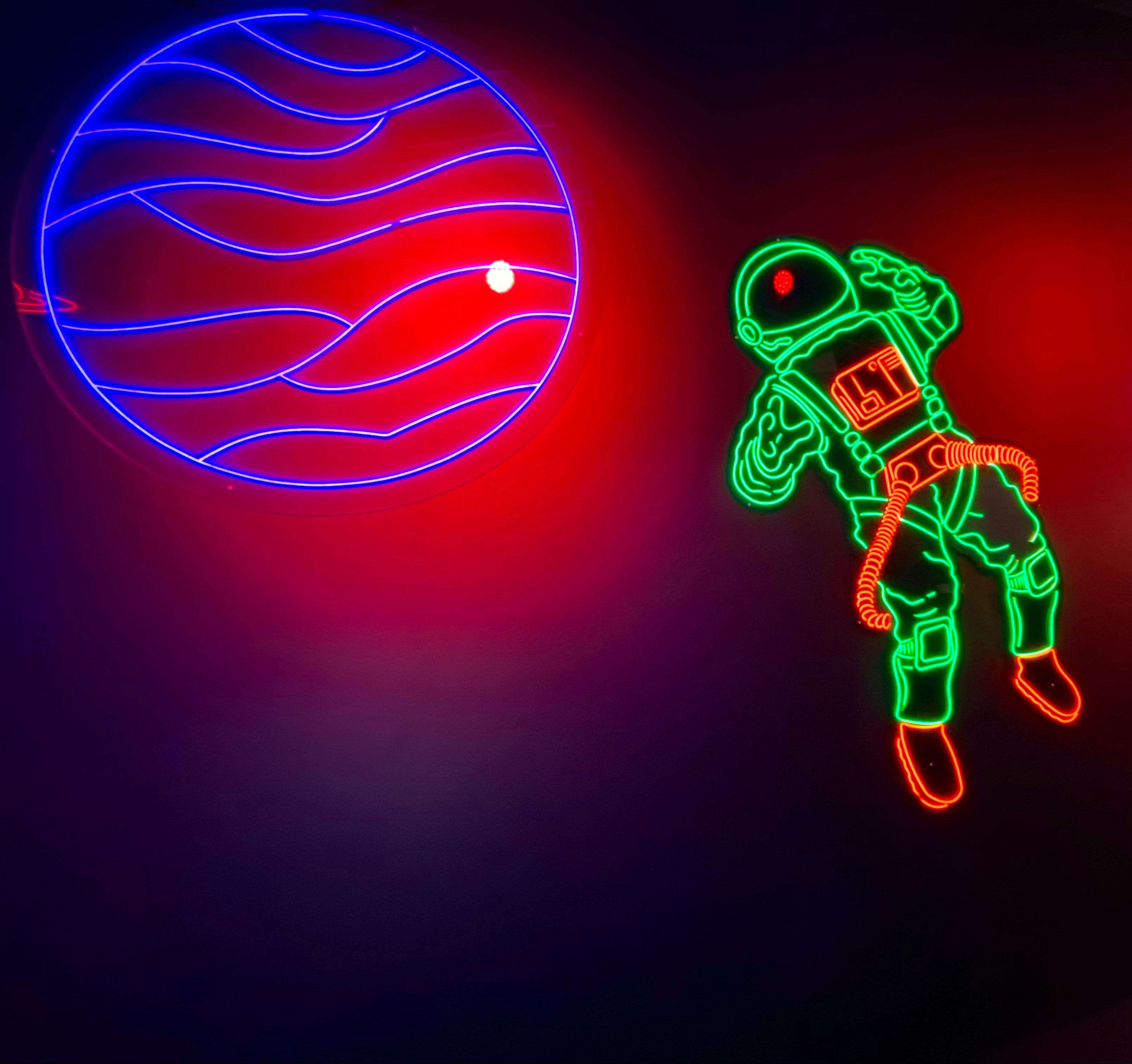 The image features a vibrant neon artwork set against a dark backdrop. On the right, a floating astronaut is outlined in bright green with orange accents highlighting the helmet visor, hoses, and boots. To the left, a stylized planet glows with blue wavy lines over a red-lit surface and a small white dot detail. The dramatic color contrast and retro-futuristic aesthetic evoke classic sci‑fi themes and neon signage design.