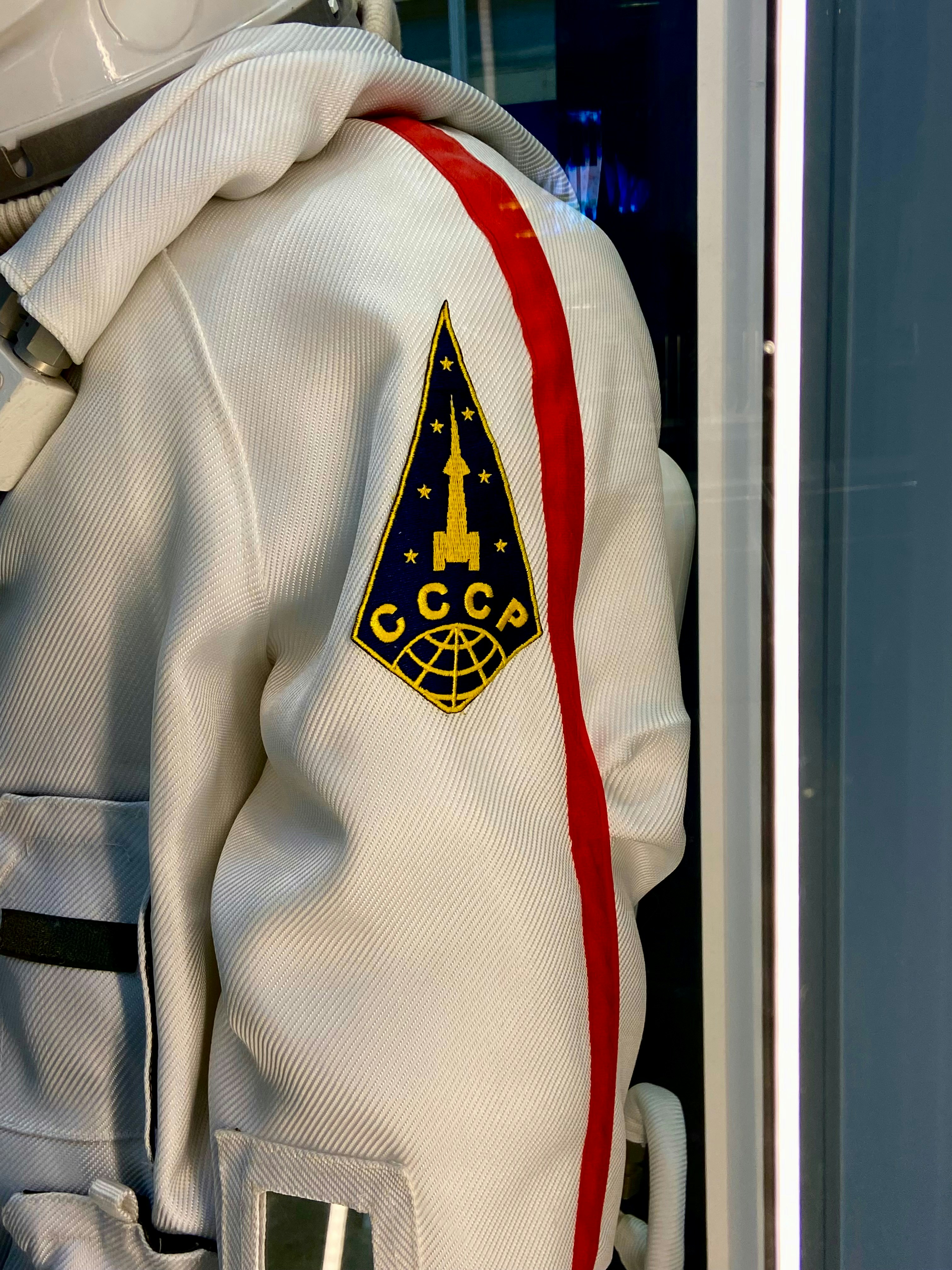 Close-up of a white space-suit sleeve with a prominent CCCP patch and red stripe, suggesting a vintage space program uniform; a hint of a helmet near the edge and a dark vertical background.