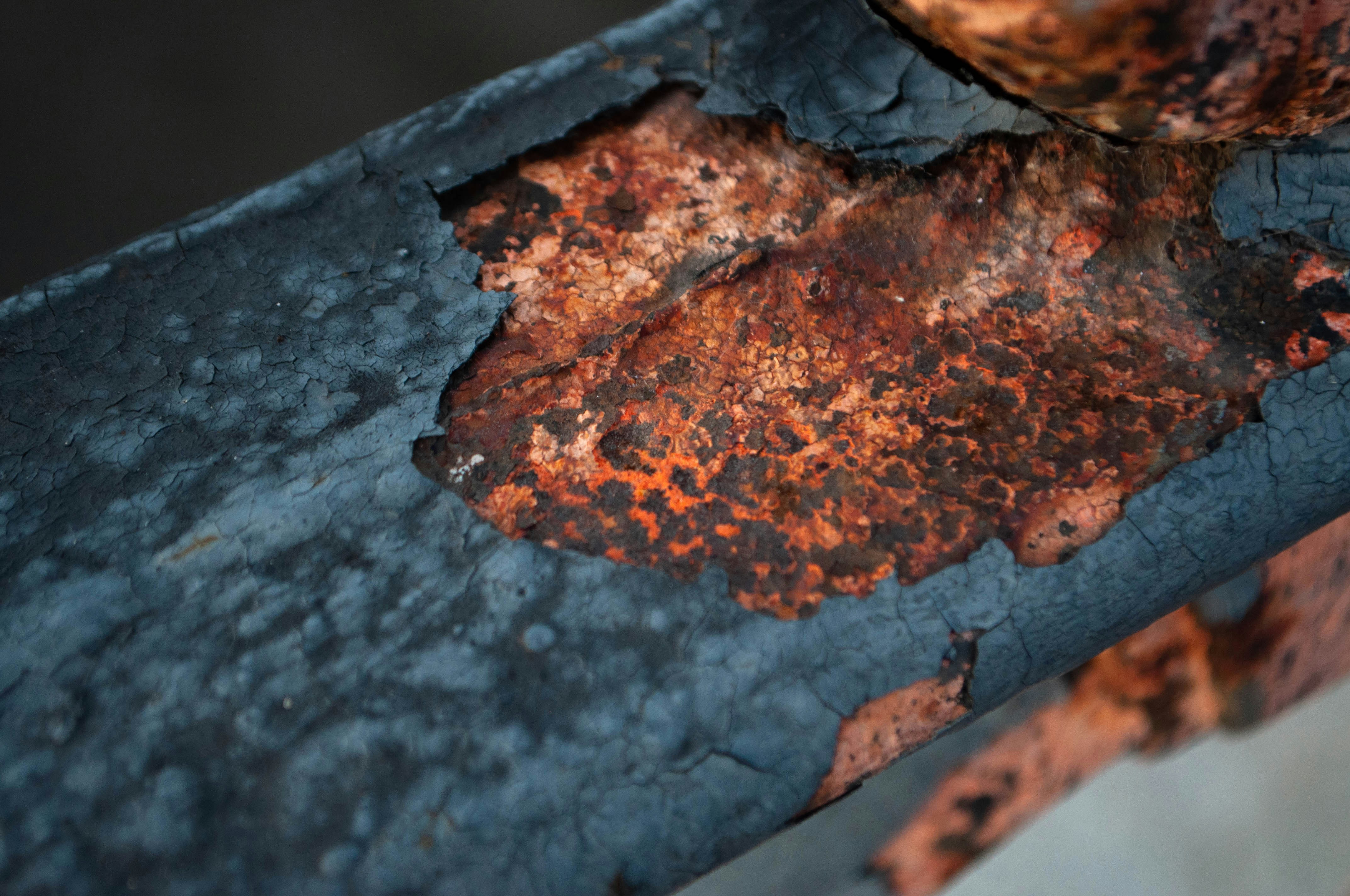 Close-up of weathered metal with peeling orange rust over a dark blue-gray surface, emphasizing texture and corrosion detail.