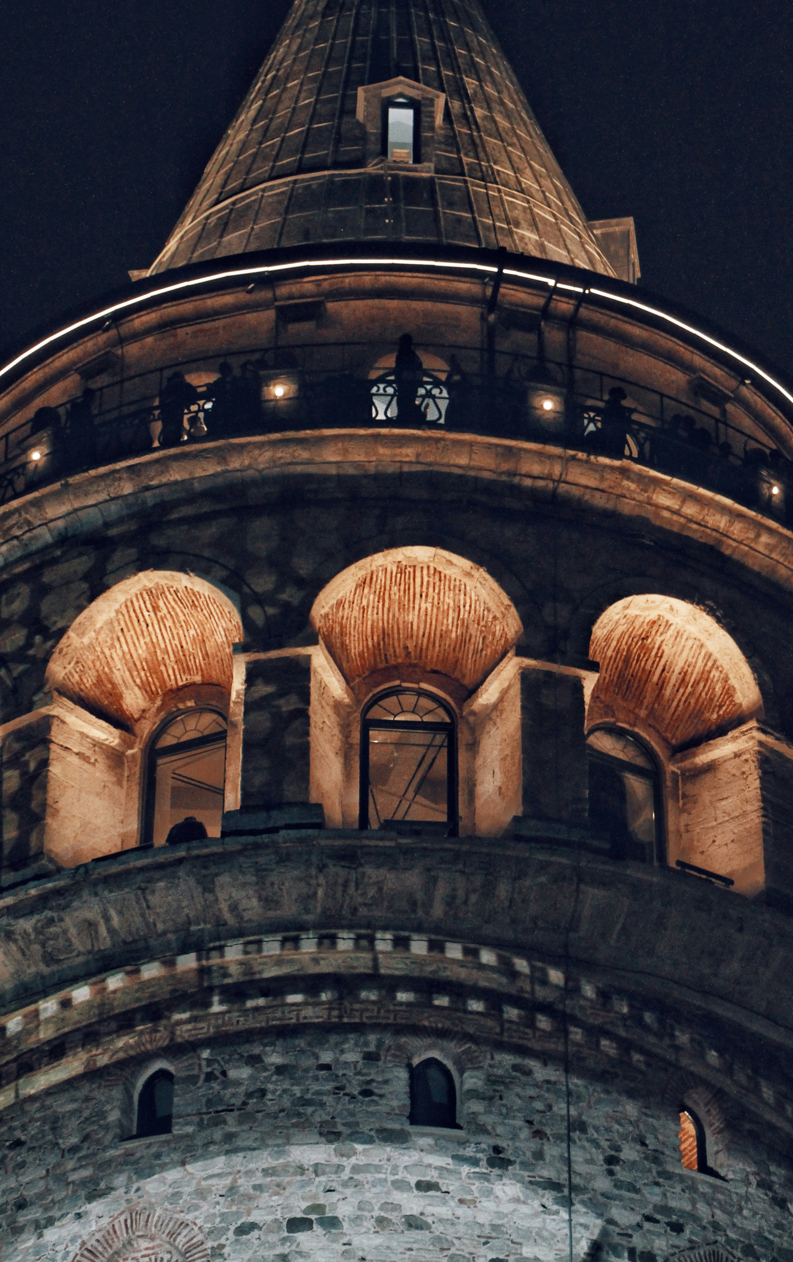 Night-time detail of a historic stone tower with glowing arches and a circular balcony, emphasizing architectural texture and warm lighting.