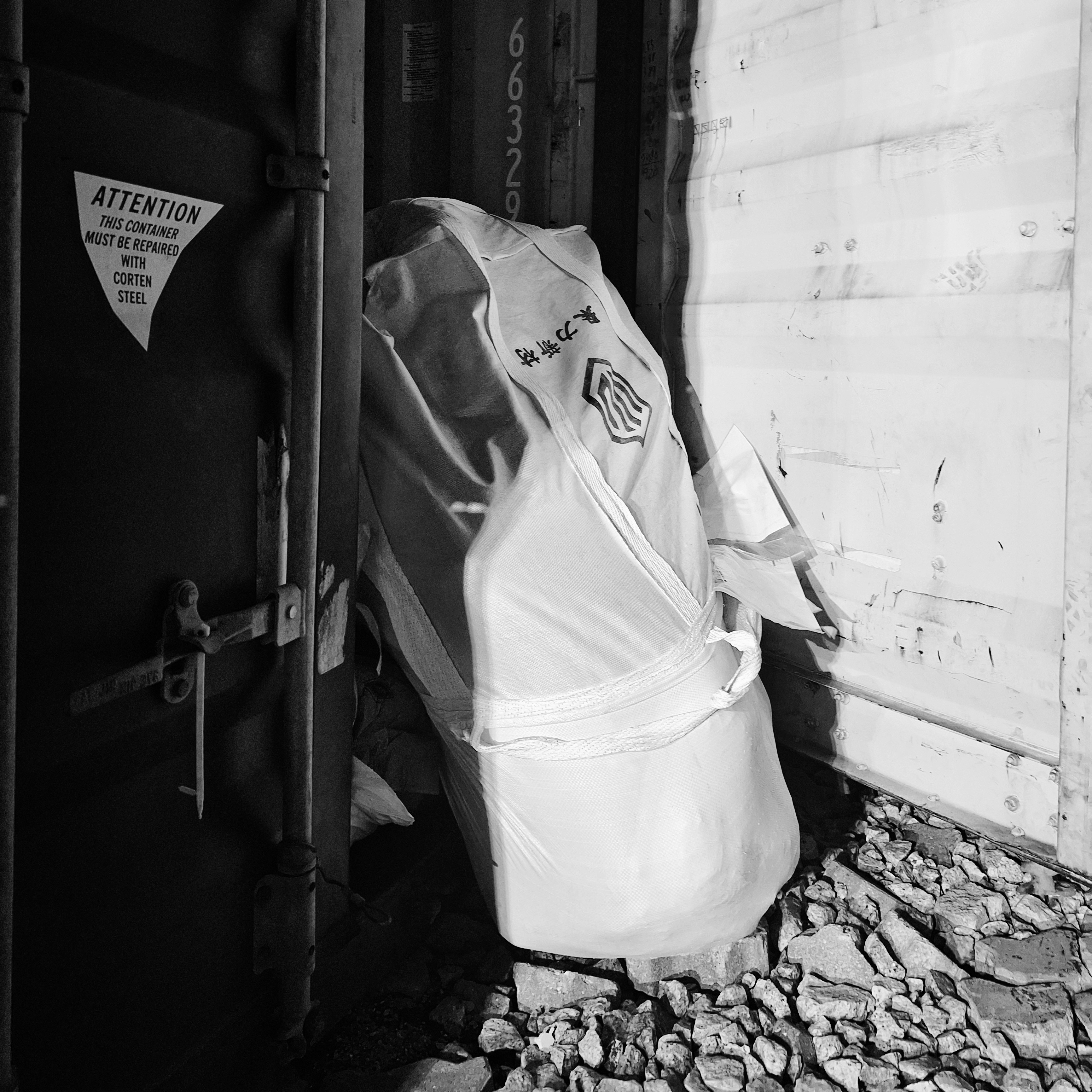 Monochrome scene of a covered, bag-laden object wedged between a dark container and a weathered rail car, resting on rocky ground. This appears to be a logistics dump or discarded cargo wrap, emphasizing texture and industrial settings.