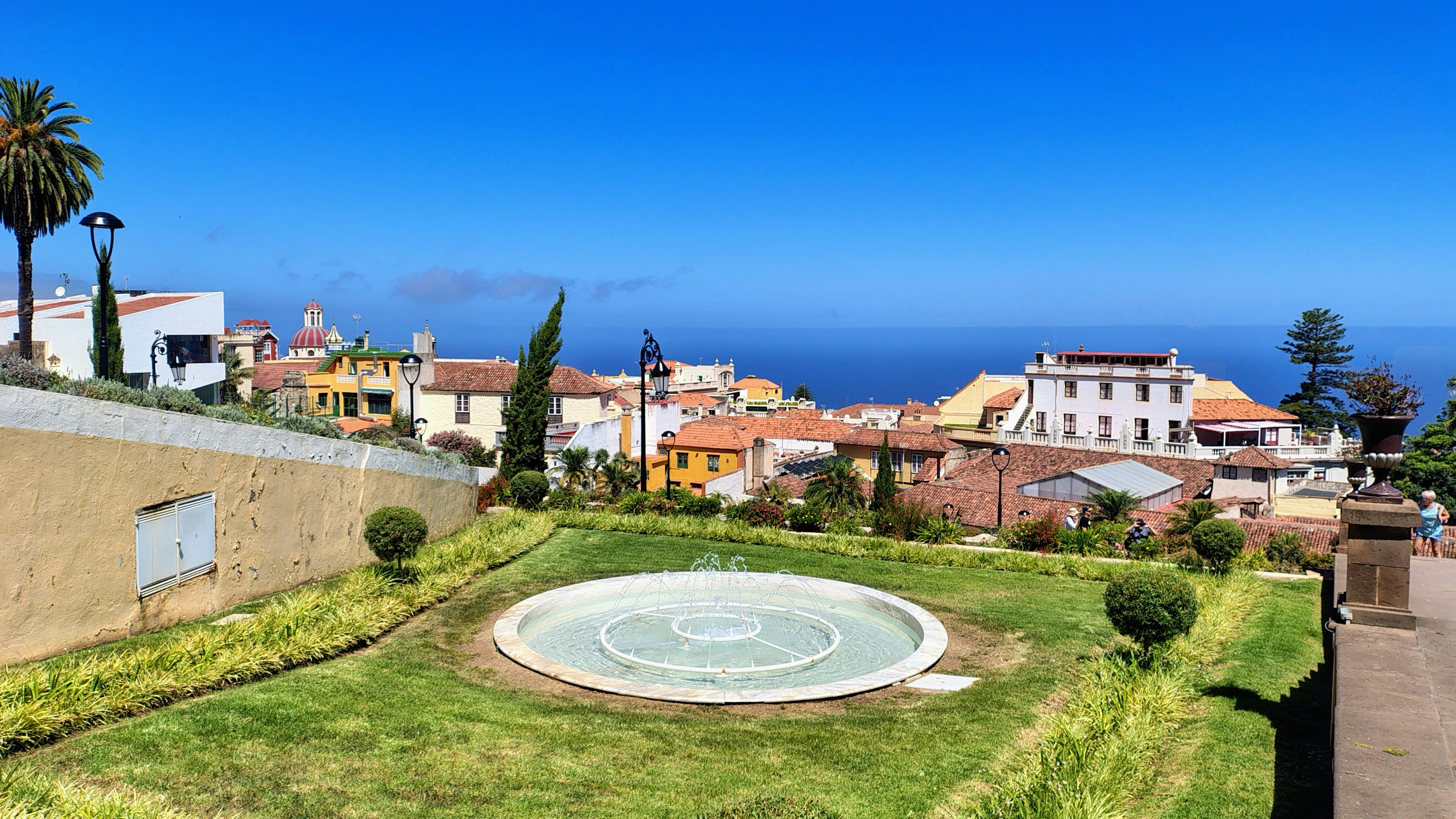Sunlit hillside plaza with orb fountain overlooking a coastal town