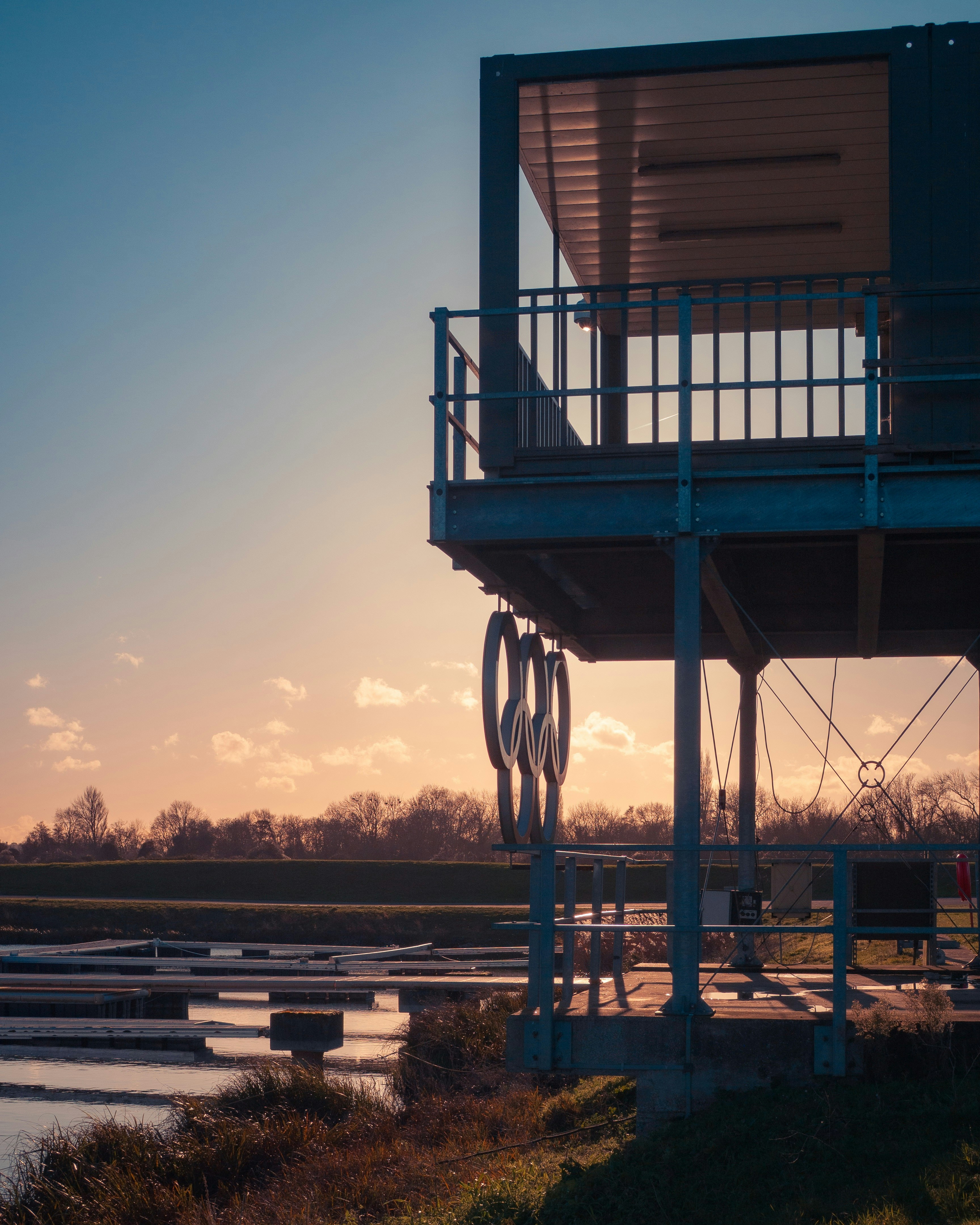 Lifebuoys hang beside a blue elevated lifeguard tower against a sunset sky, with a calm river and distant trees in the background.