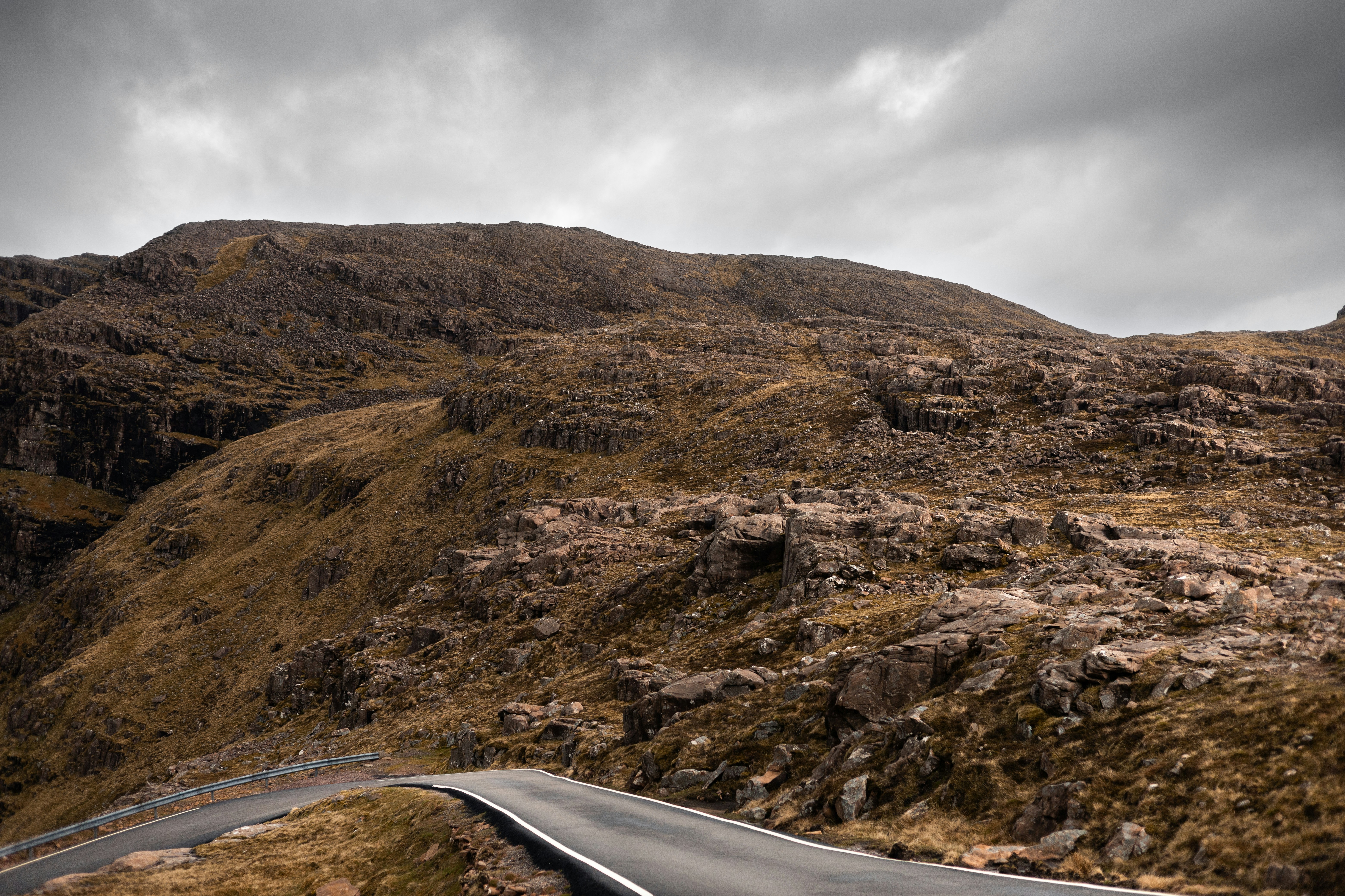 A narrow, winding mountain road curves through a rugged, dark rocky landscape beneath a dramatic, storm-clouded sky.