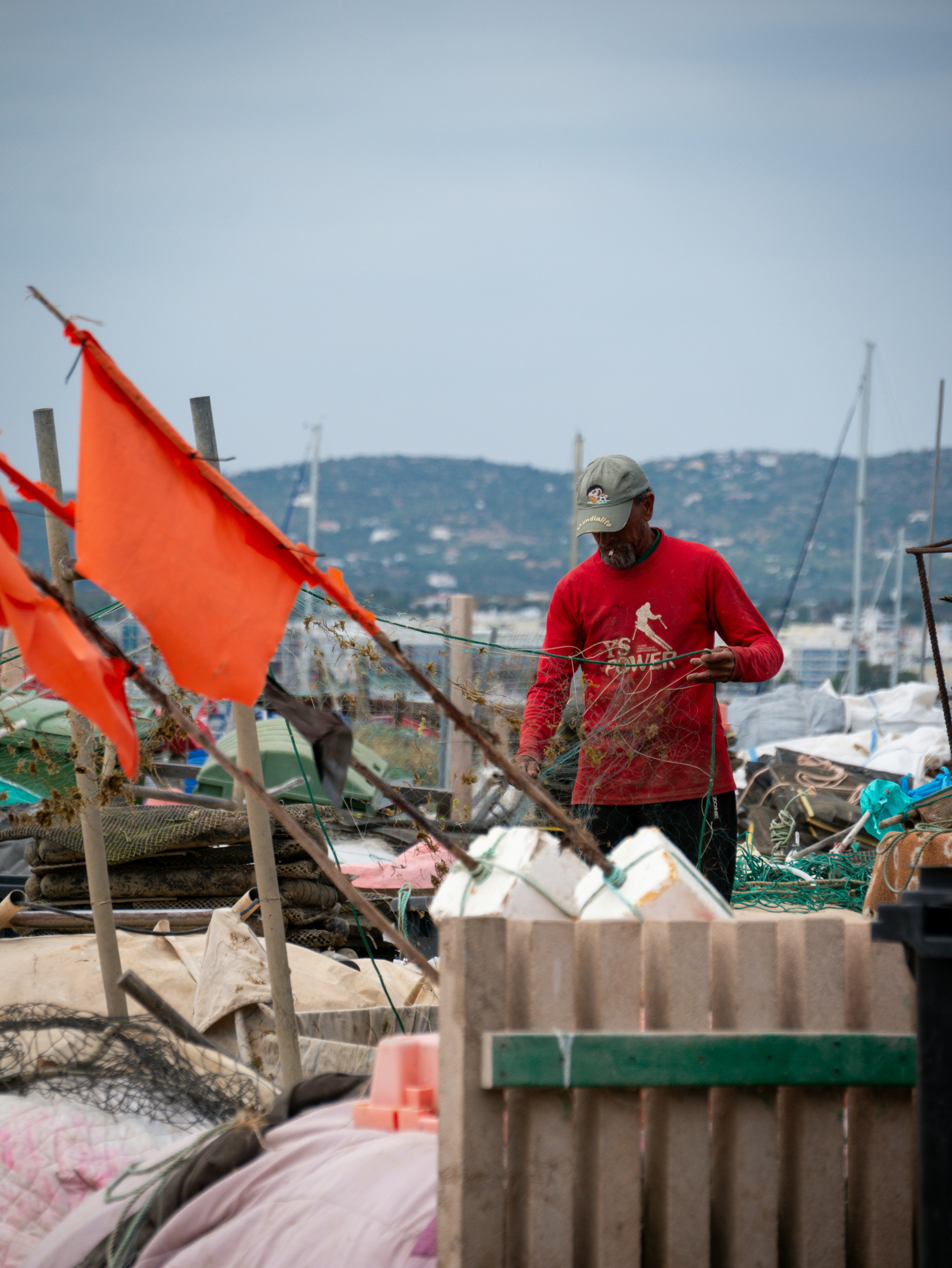 A man in a red shirt stands among clutter and nets at a harbor, with orange flags and distant hills in the background.
