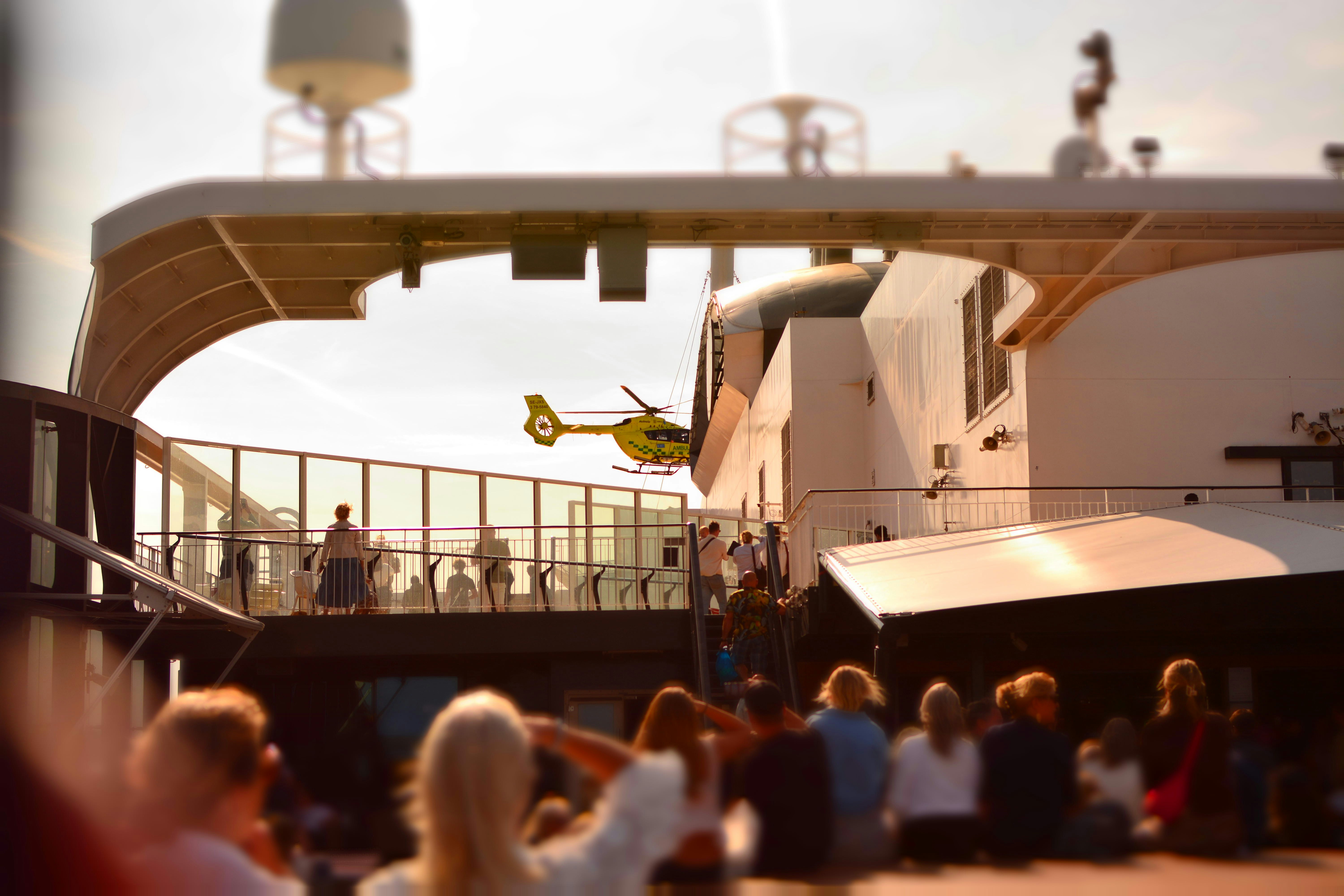 Crowd seated on a ship deck watches a helicopter hovering near the rail, with a glass-walled observation walkway and modern architecture in the background.