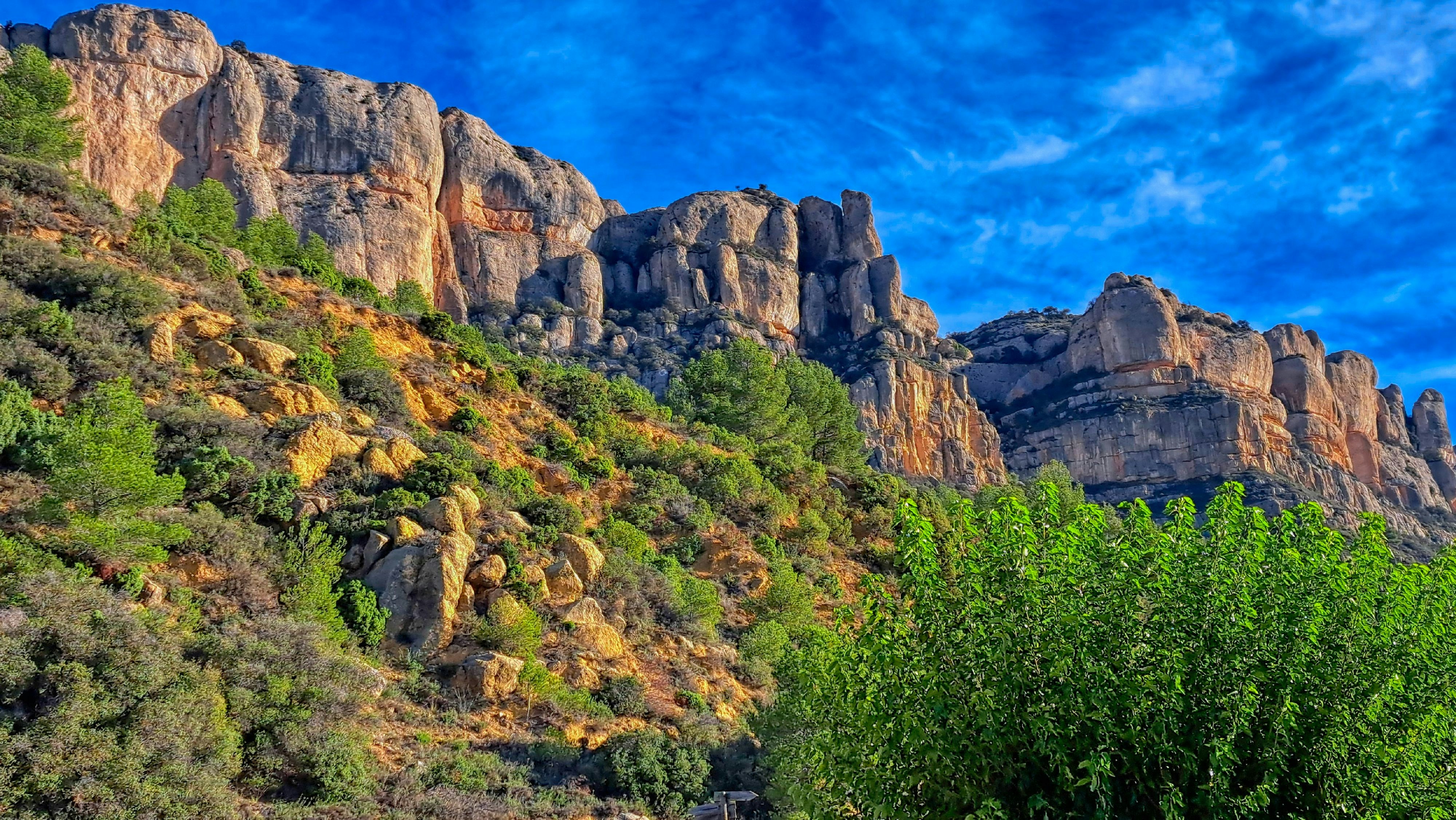 Rocky cliffs rise above evergreen slopes under a vivid blue sky, with sunlight illuminating the rugged terrain and trees along the hillside.