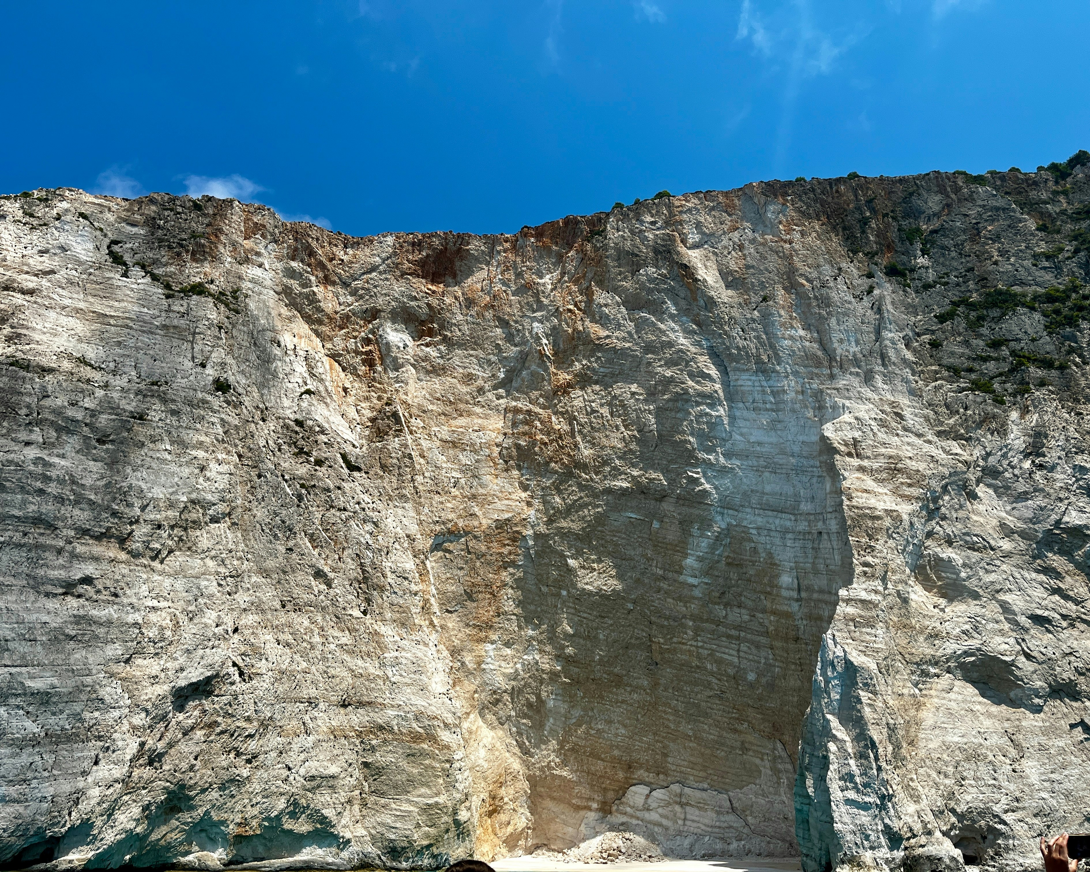 Rusty cliff face rises above a narrow sandy beach with a turquoise sea at the base; a vivid blue sky crowns the scene and a carved arch-like rock formation dominates the center.