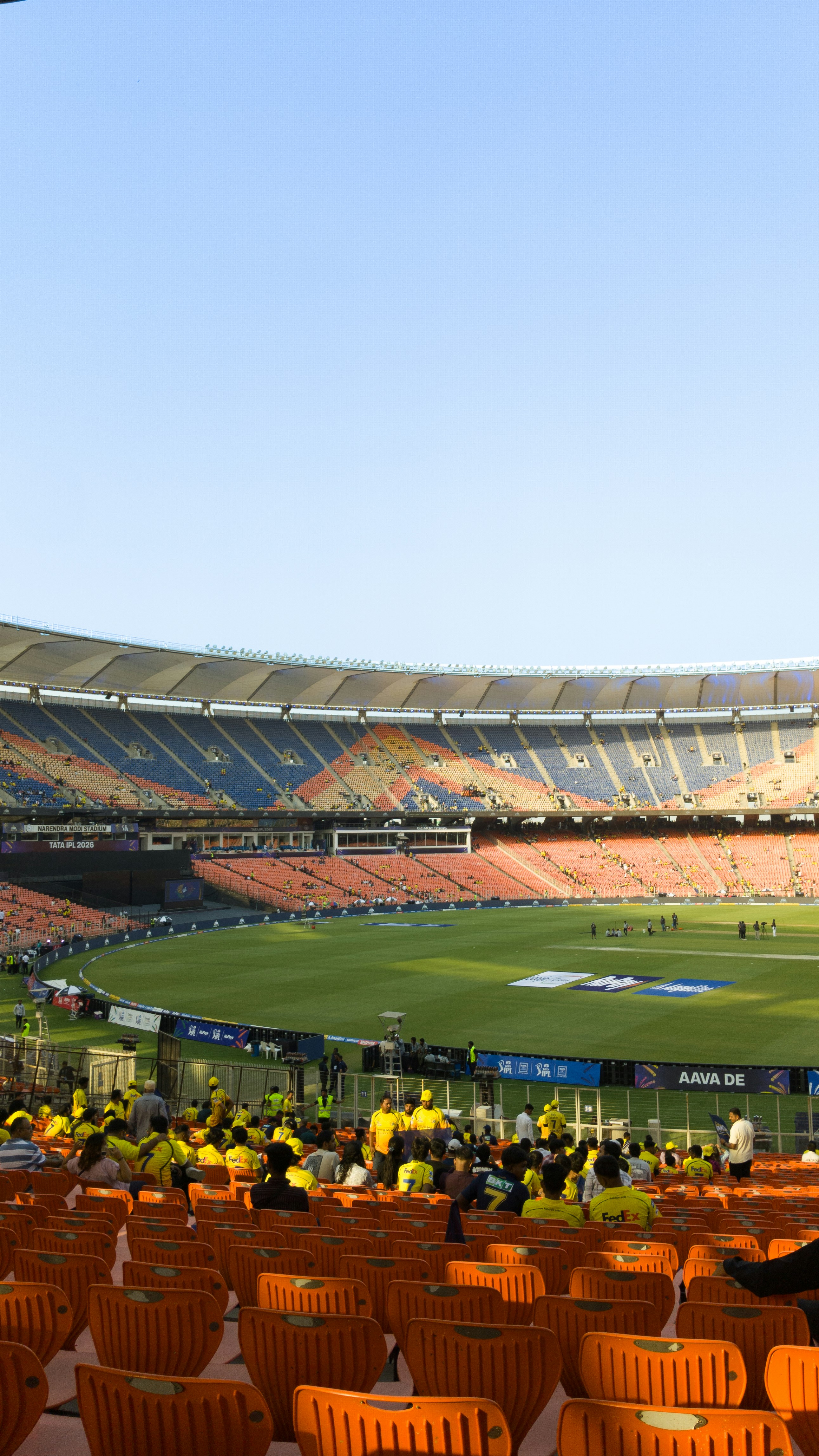 A cricket stadium with orange seating and a green field under a clear sky