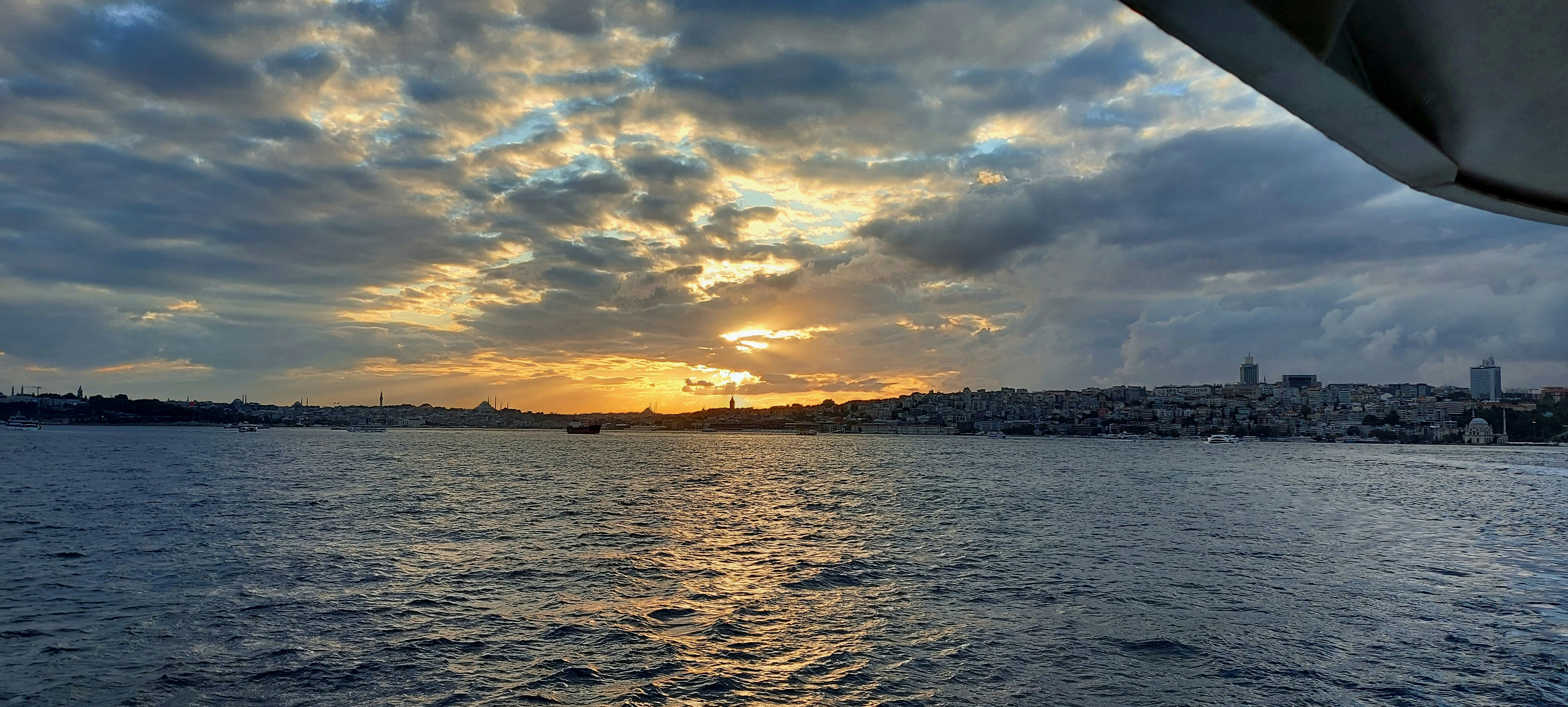 Sunset over a calm bay with scattered clouds and distant city silhouette; water reflects the warm sky, captured from a low vantage near the shore.