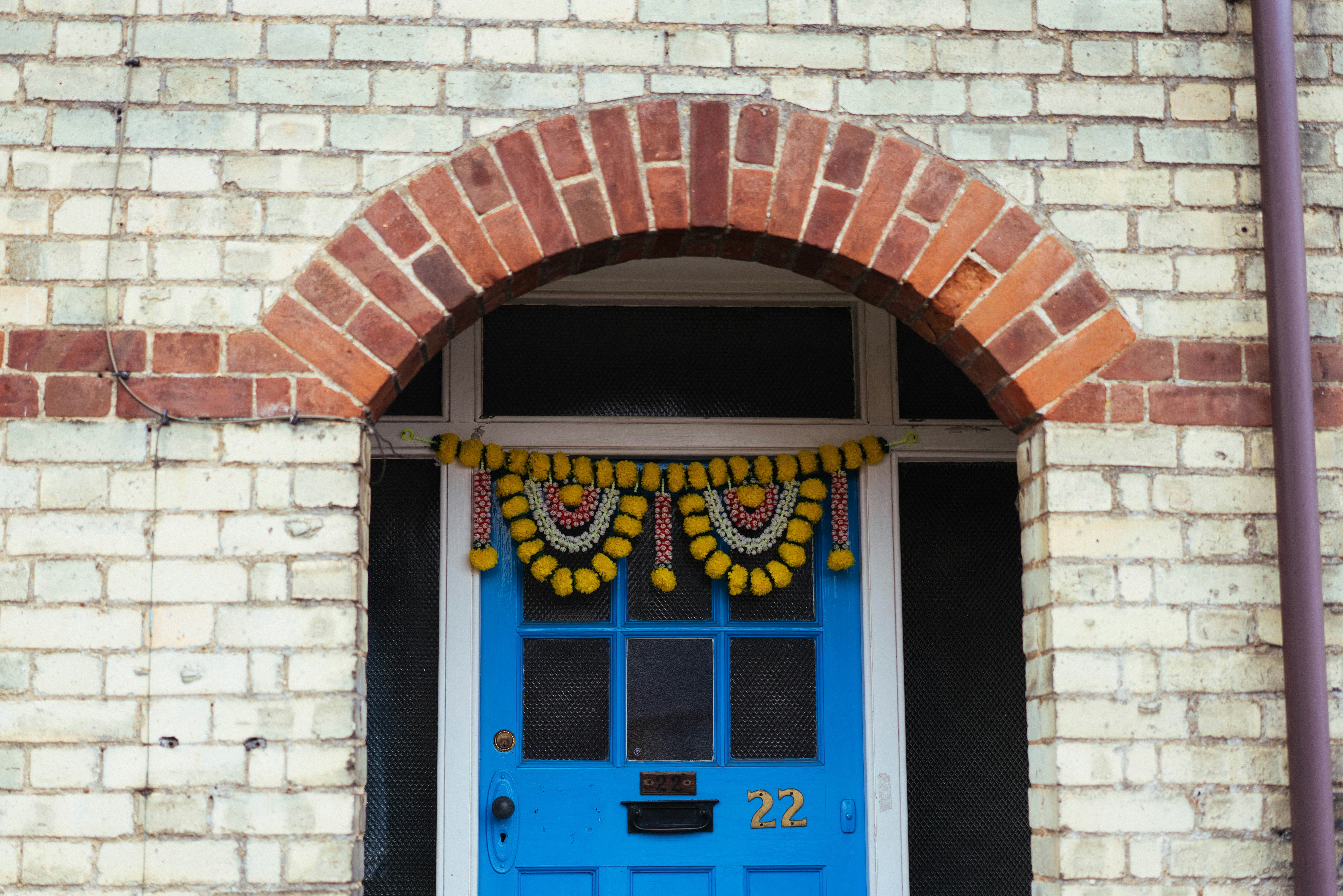 Bright blue front door framed by a brick arch with decorative garlands, set in a pale brick facade; a small mailbox and house number 22 visible on the door. Ominously, the frame hints at a doorway rather than a broader street scene.