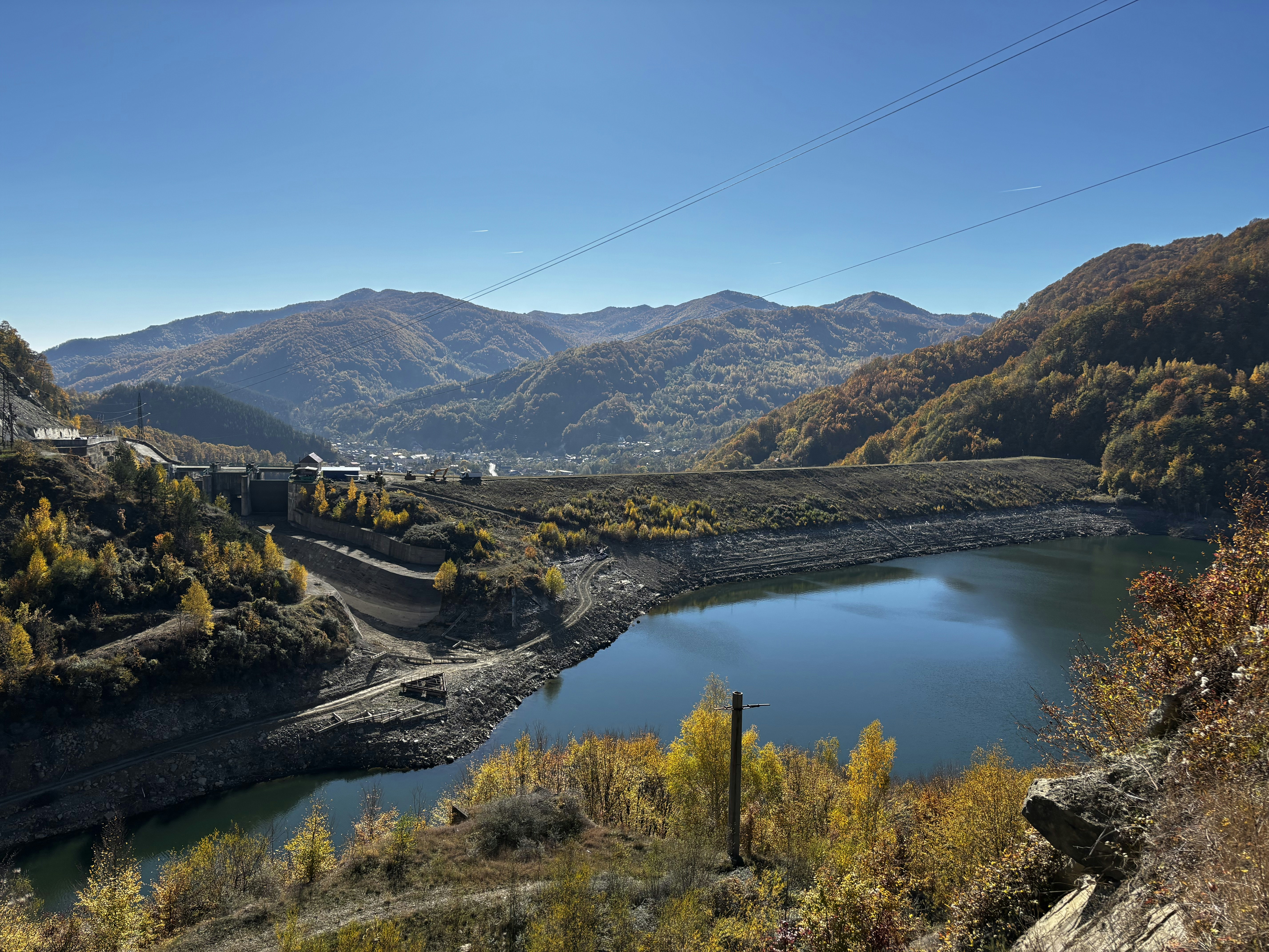 Autumnal valley with a calm reservoir edged by rocky banks and yellow foliage, set against distant forested hills and a clear blue sky.