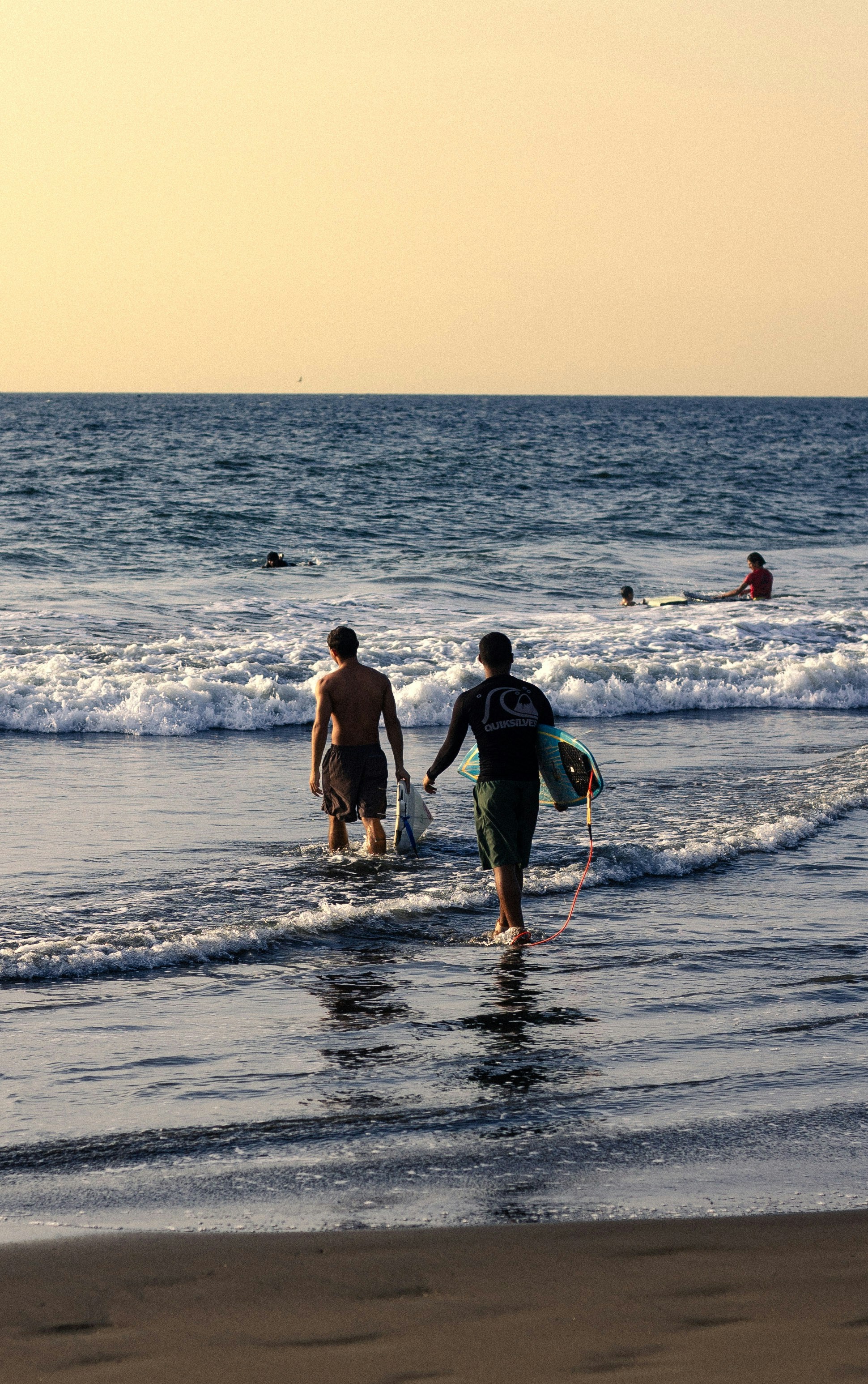 Two surfers return to shore as ocean waves roll in under a warm coastal glow