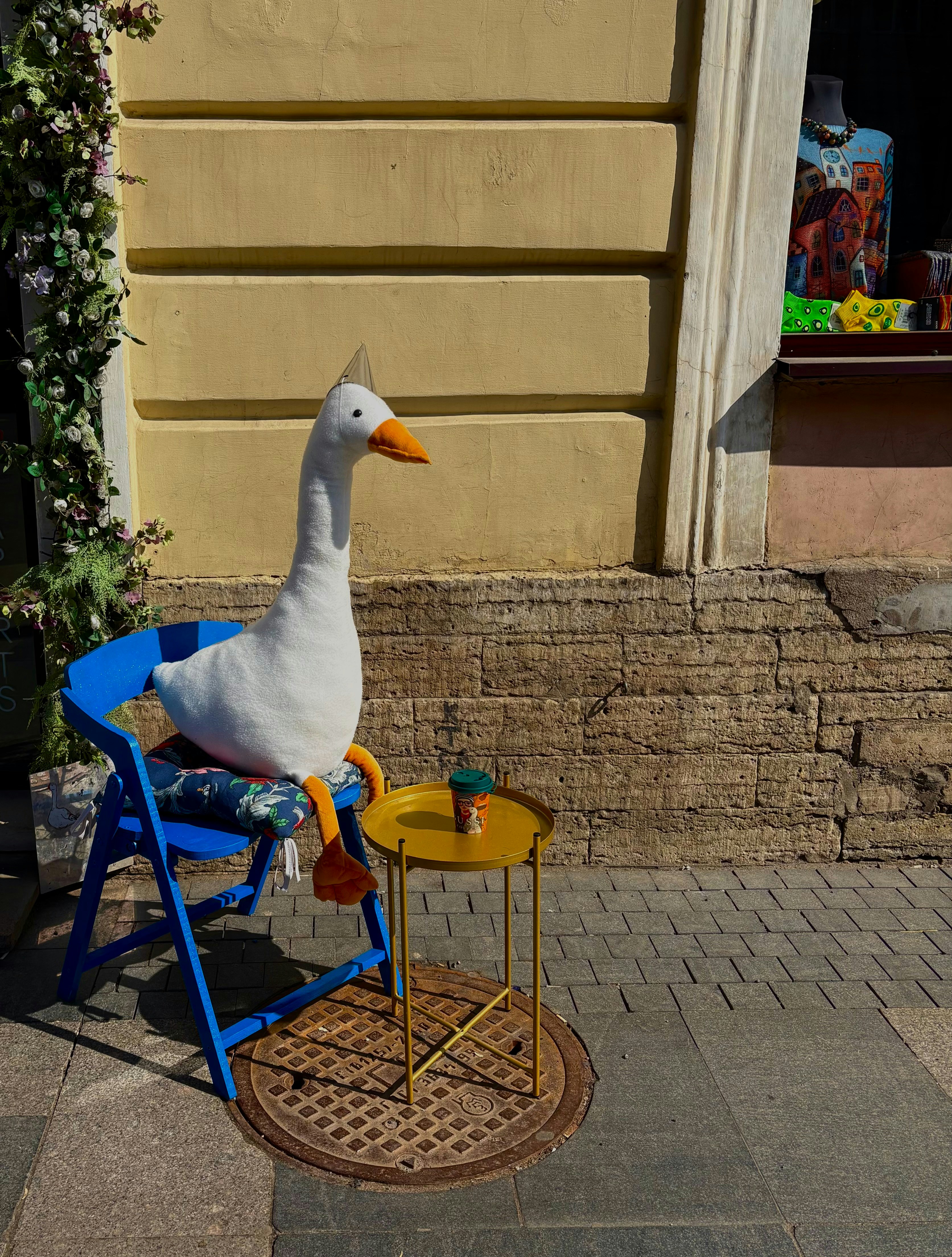 The image features a large white goose sculpture seated on a bright blue folding chair, posed as if enjoying a quiet moment. In front of it stands a small yellow metal side table holding a disposable coffee cup. The setup rests atop a circular manhole cover on a paved sidewalk. Behind the scene, a pale yellow wall with textured panels meets a rustic stone base, while a window display with colorful decor and a trailing floral arrangement add lively details to the urban tableau.